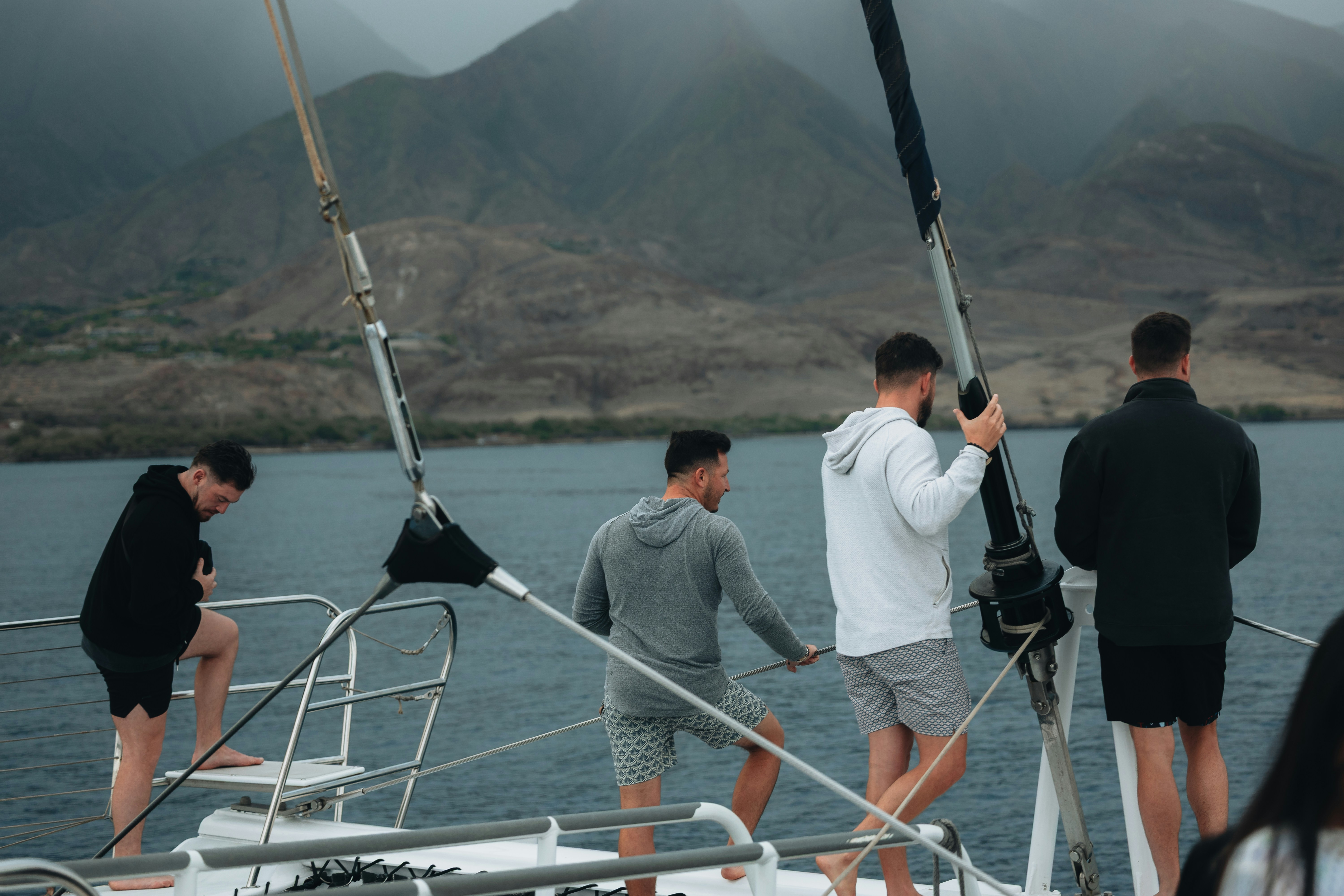 Four men on a sailboat adjust lines against a mountainous coast and calm water backdrop.