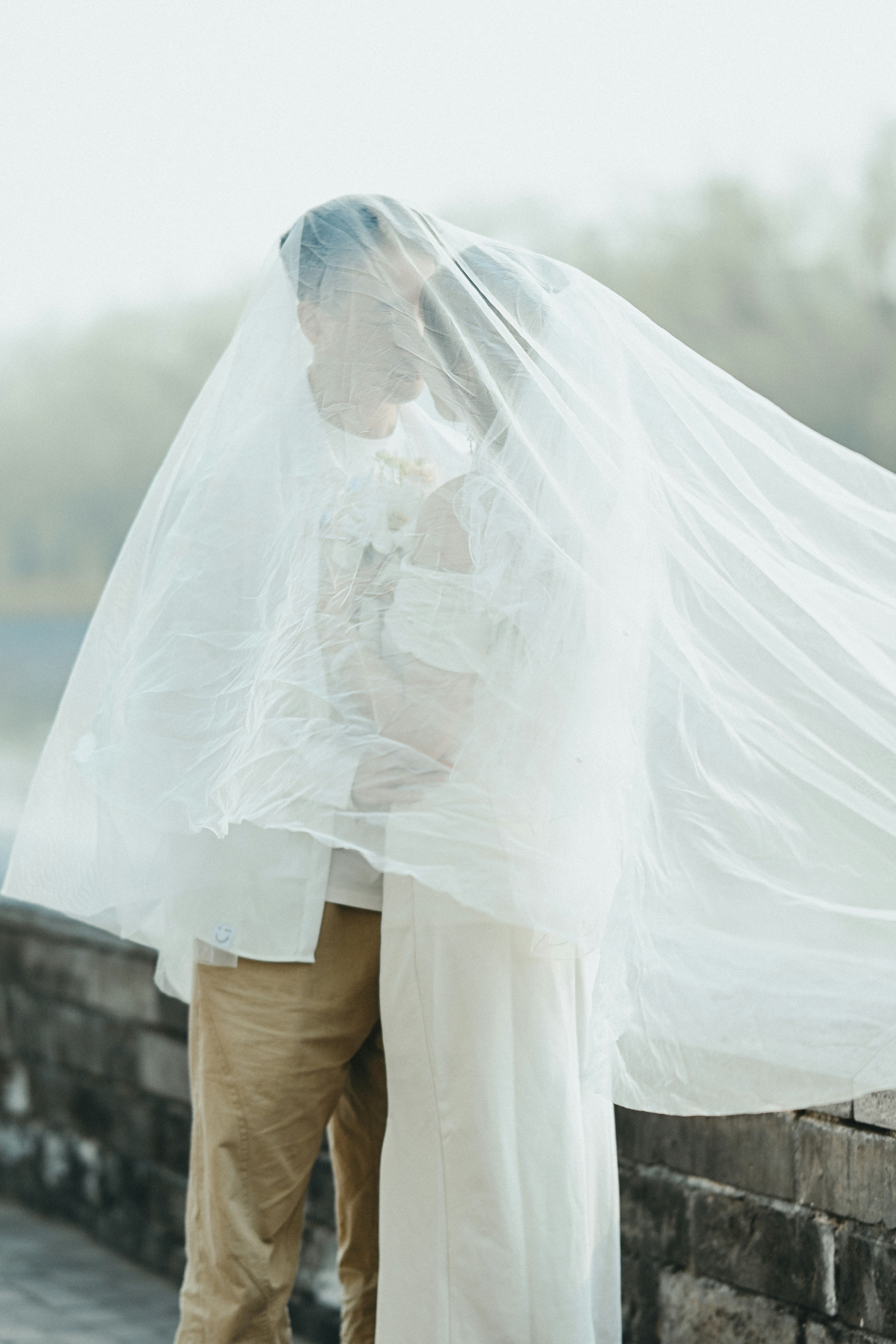 Veil of Quiet: A Bride and Groom Embrace Under a Flowing Sheer