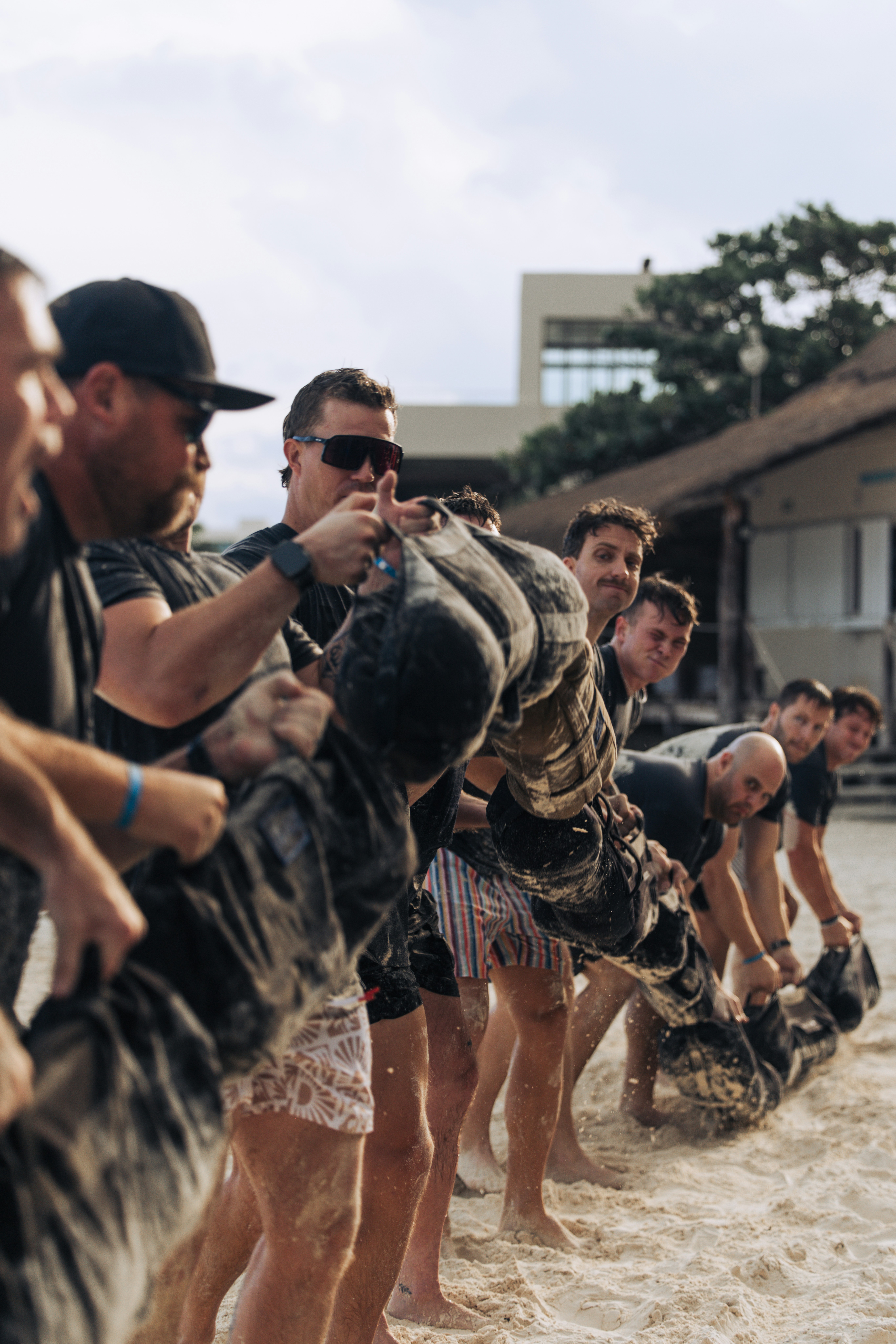 Group of men pulling a rope together on sandy beach, mid-action, with onlookers in the background; candid team activity shot at a sunny seaside location. Photograph is a documentary capture of a sport or challenge event.