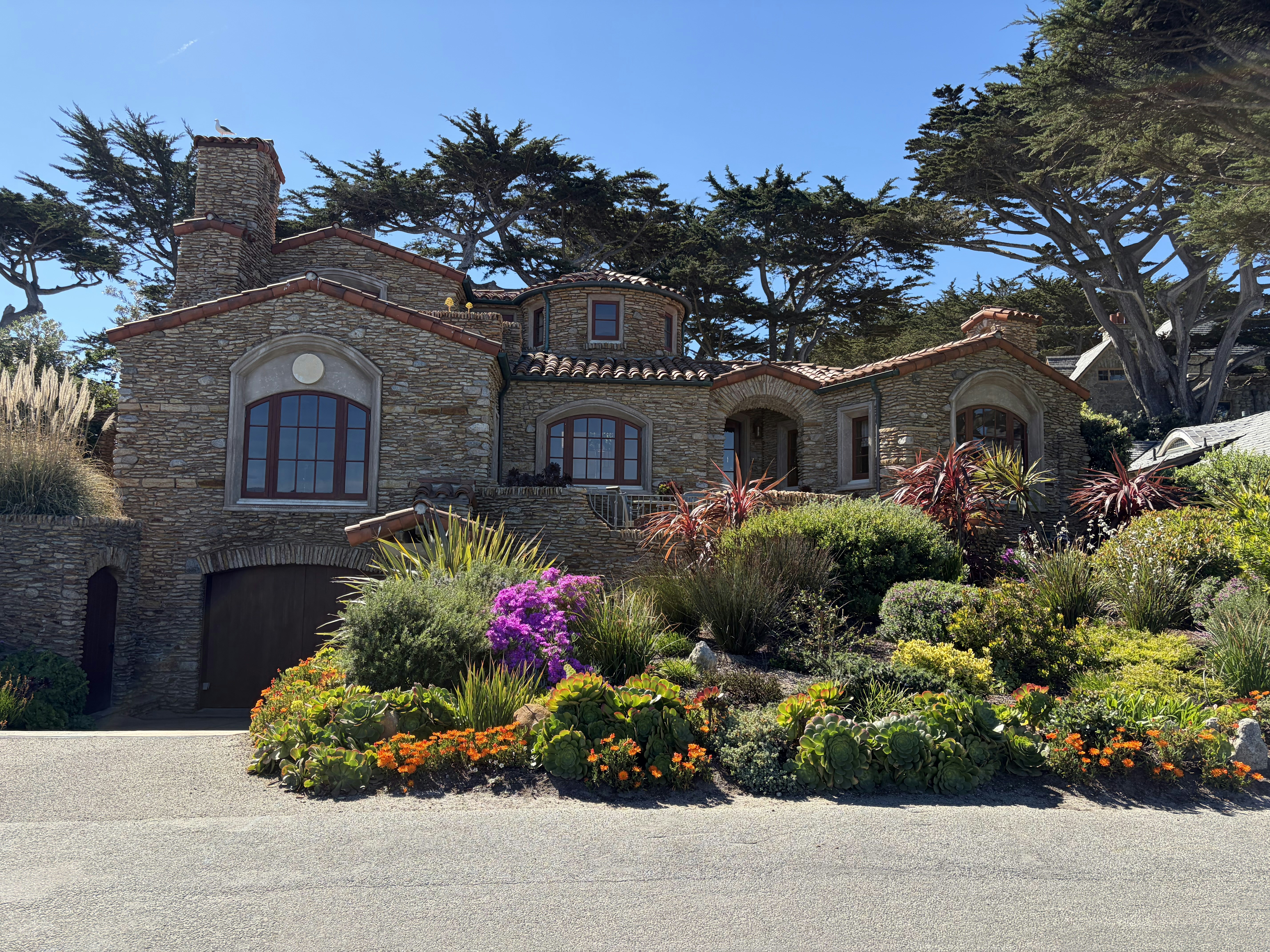 Colorful flower beds frame a stone hillside house under a bright blue sky, with lush landscaping and a gravel driveway; a picturesque residential scene.