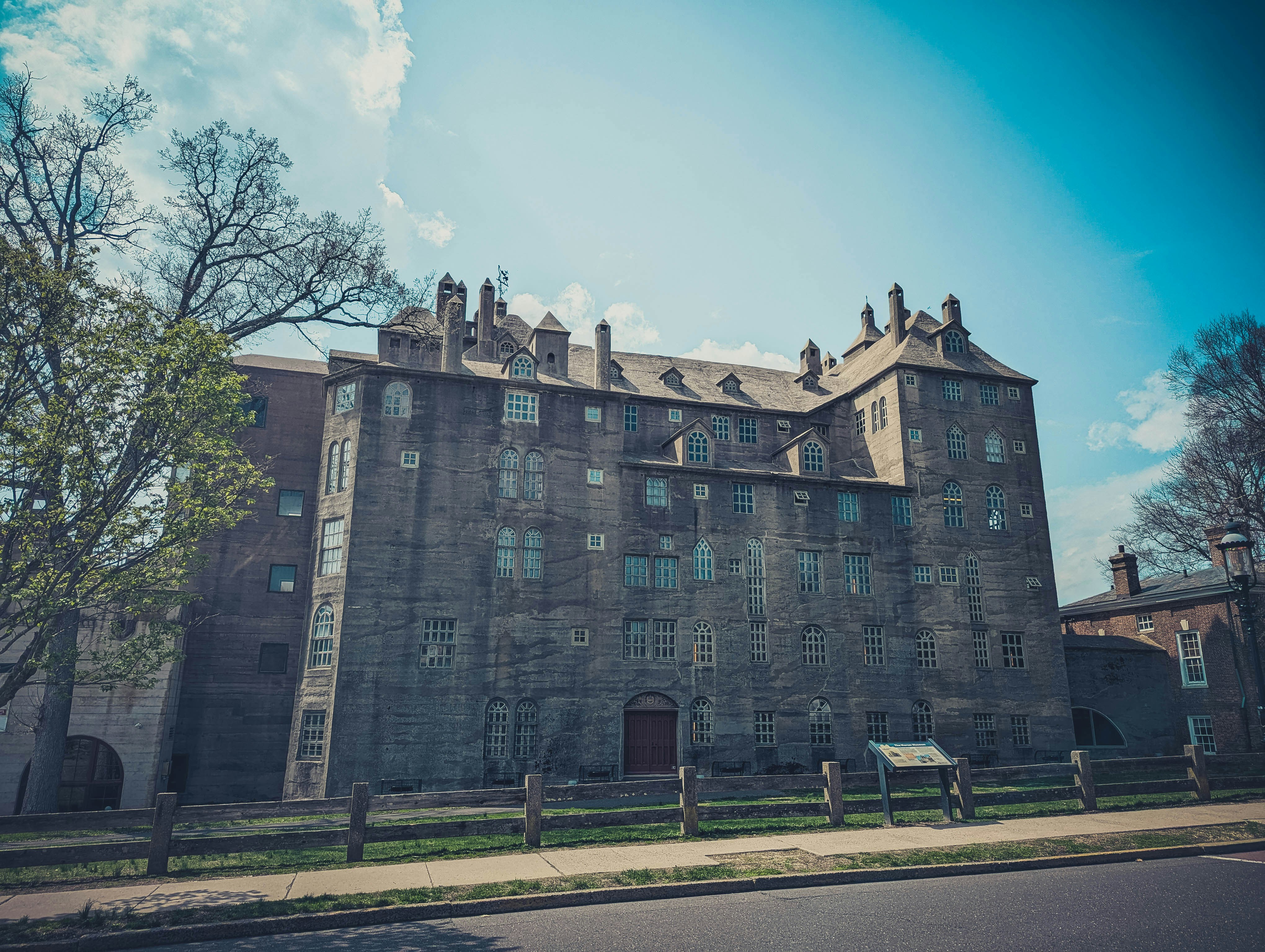 A historic stone building sits along a quiet street with a wooden fence and trees framing the façade, photographed in daylight. The image documents architectural details and mood of an old fortress-like structure.