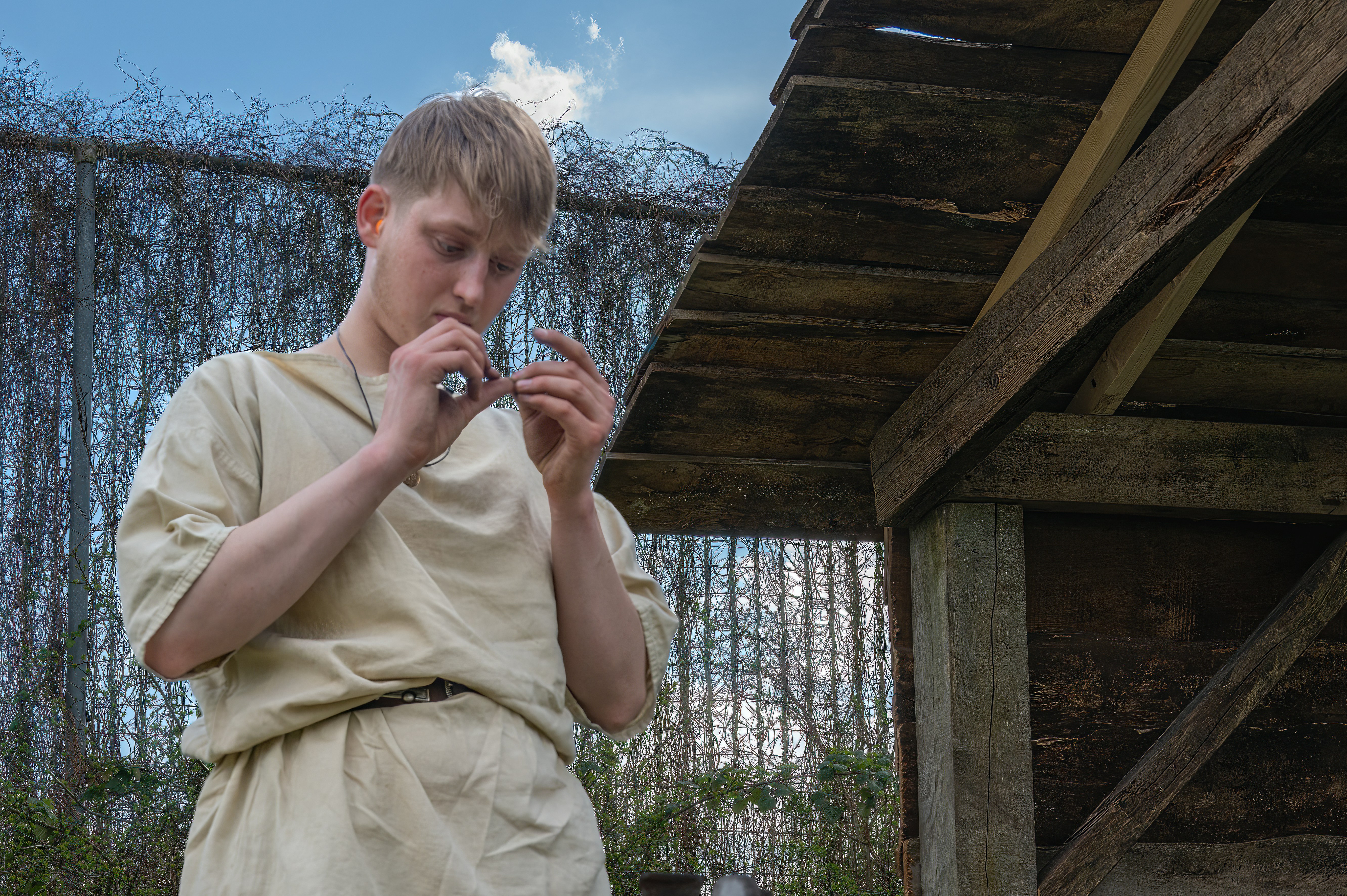 Young man in simple tunic adjusting a small tool near a wooden shelter