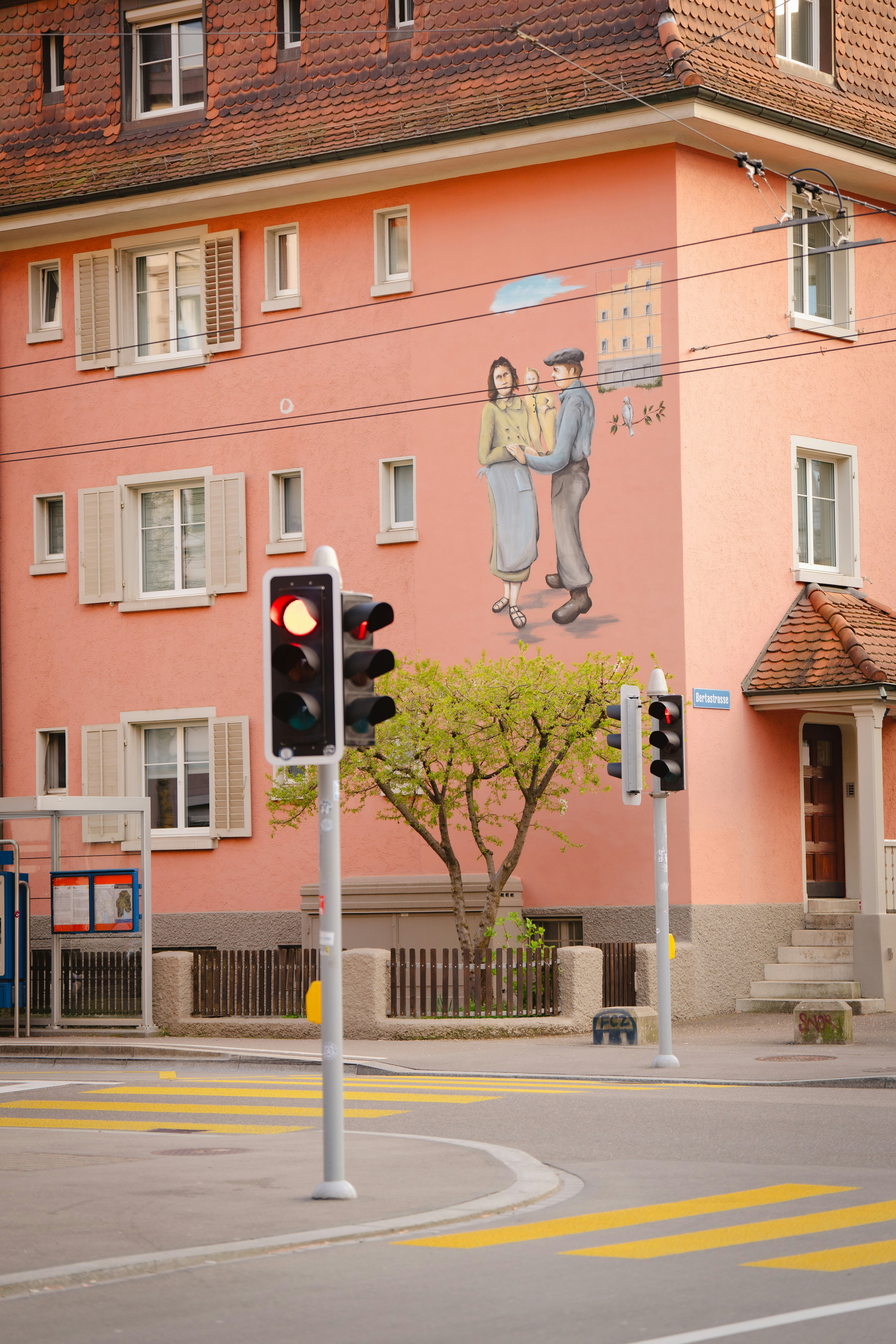 Colorful mural on a peach apartment building visible beside a traffic intersection; foreground shows street signals, crosswalk lines, and a small tree, with a calm urban scene beyond.