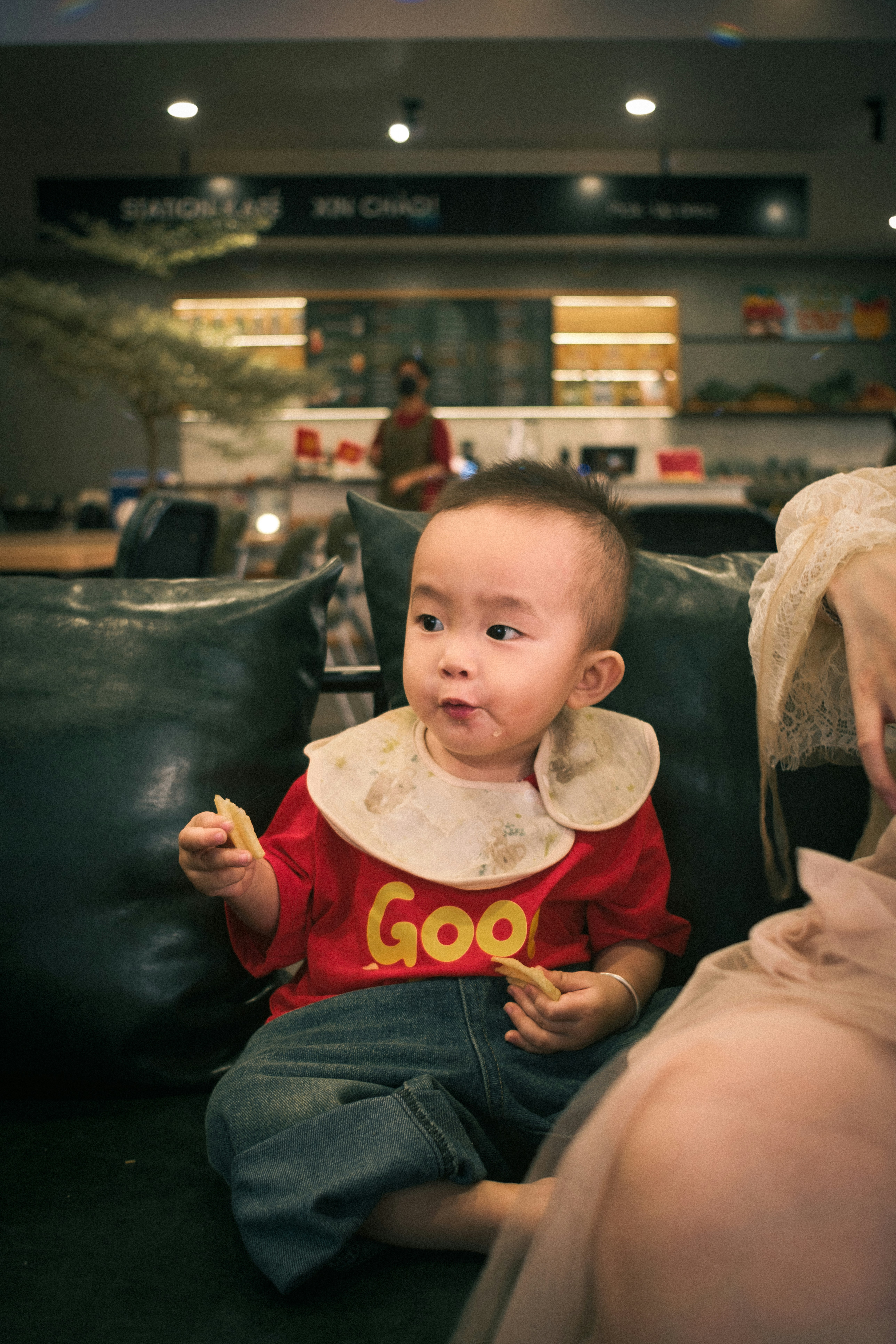 Baby in a Red Shirt sampling snacks on a cozy couch inside a cafe