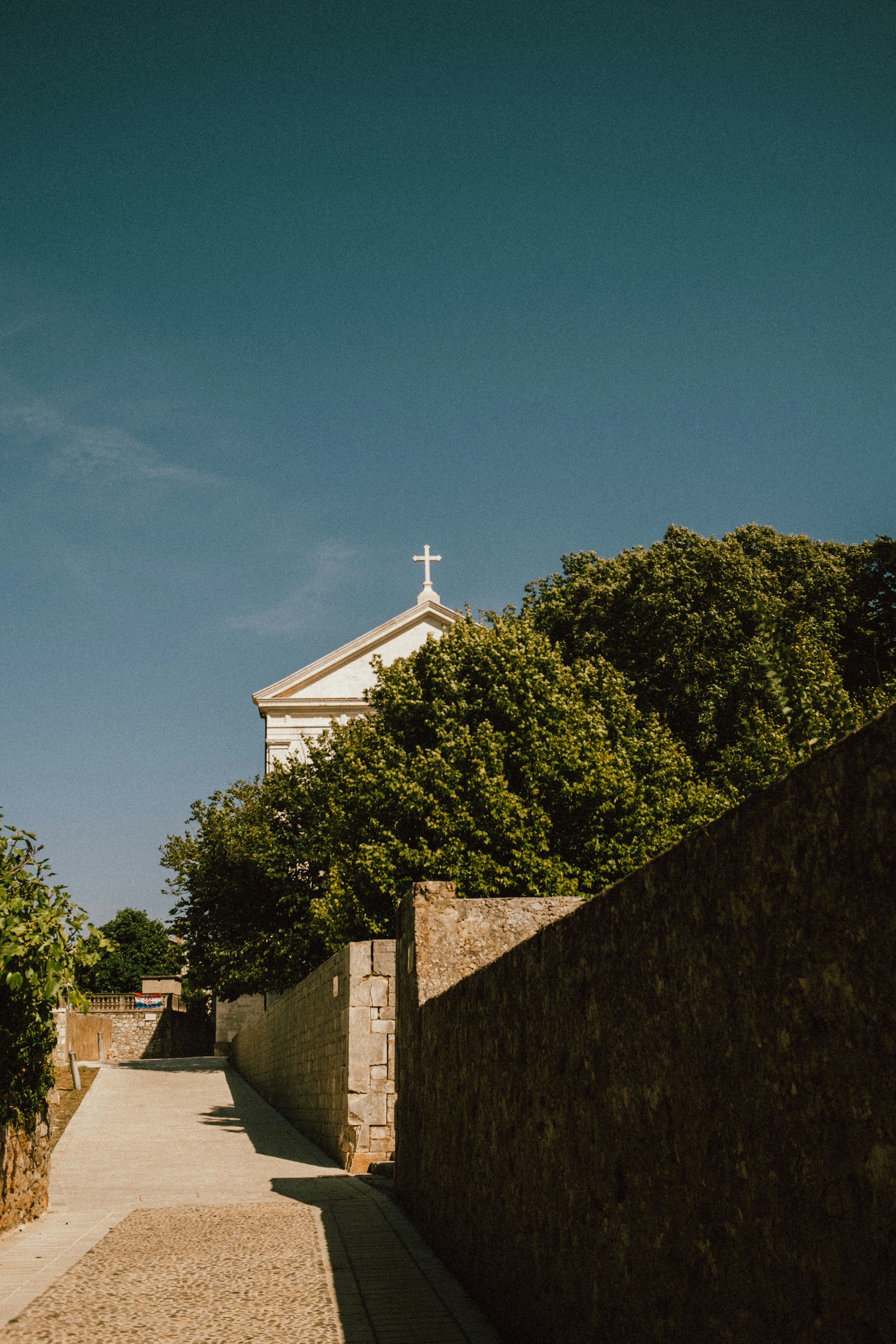 Cross atop a quiet stone path: church facade peeking through a canopy of trees