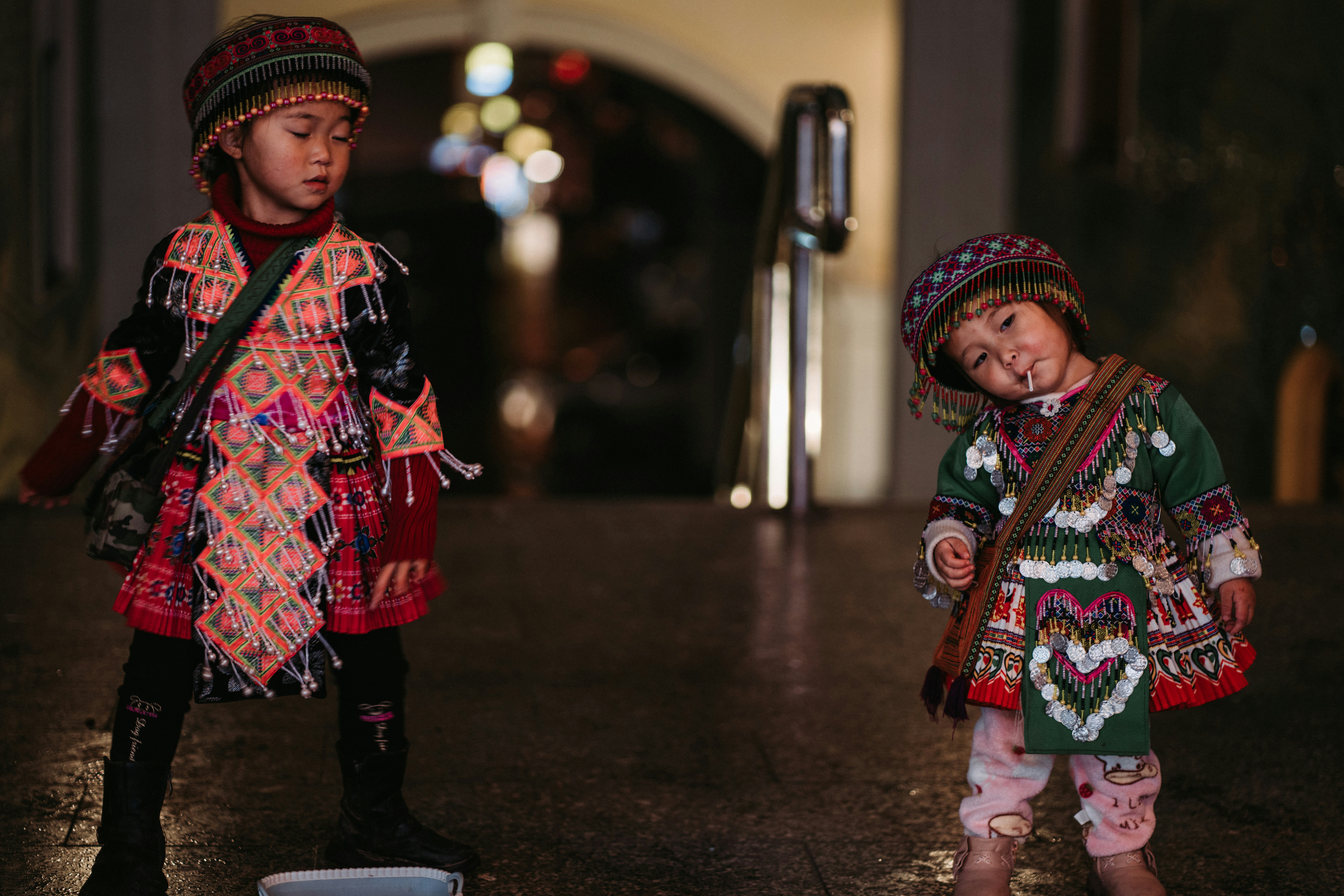 Two children in vibrant traditional attire stand on a dim street at night