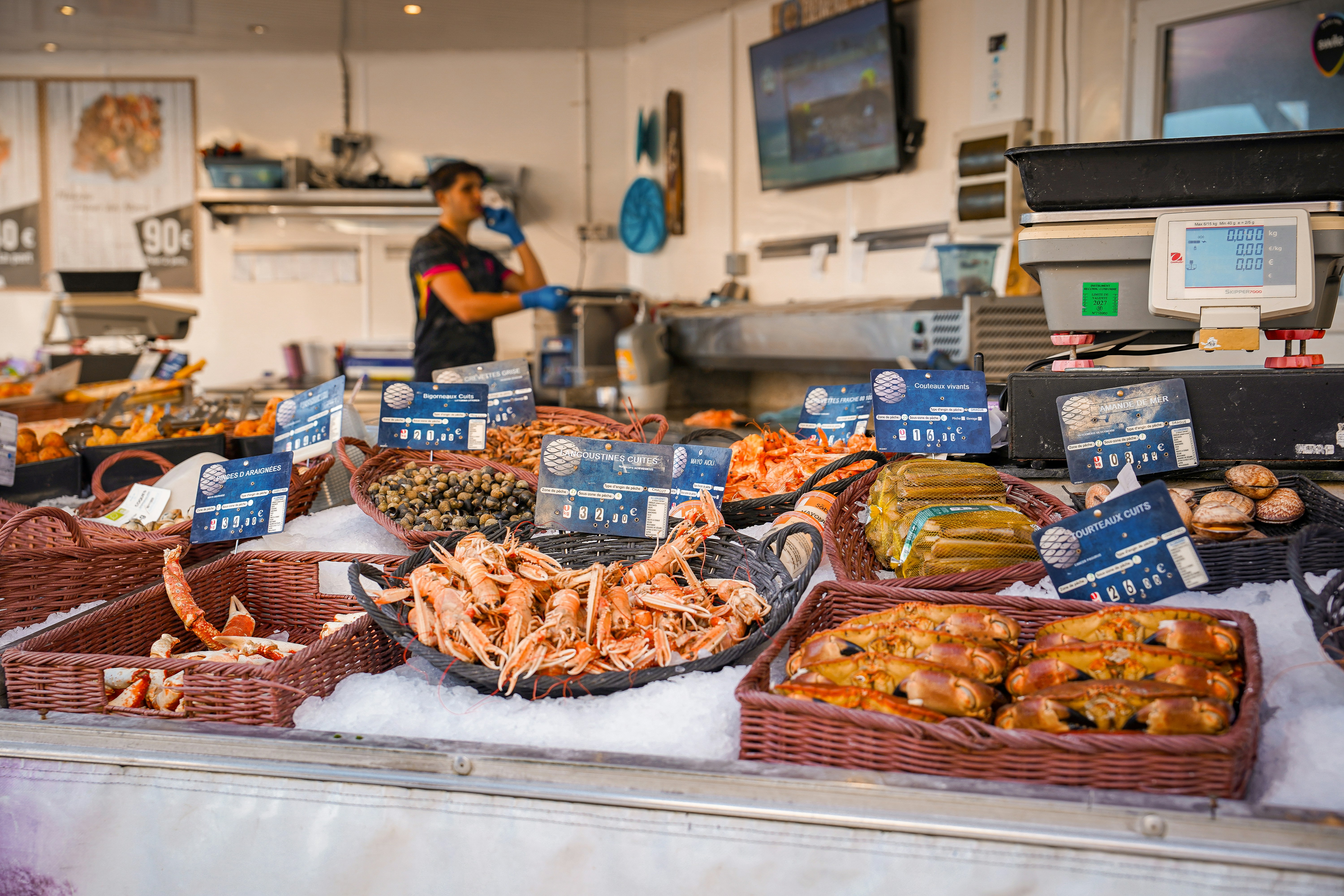 Seafood market display with shrimp, crabs, and other shellfish on ice; scale shown by a vendor in the background. A retail environment focused on fresh produce and seafood signage.