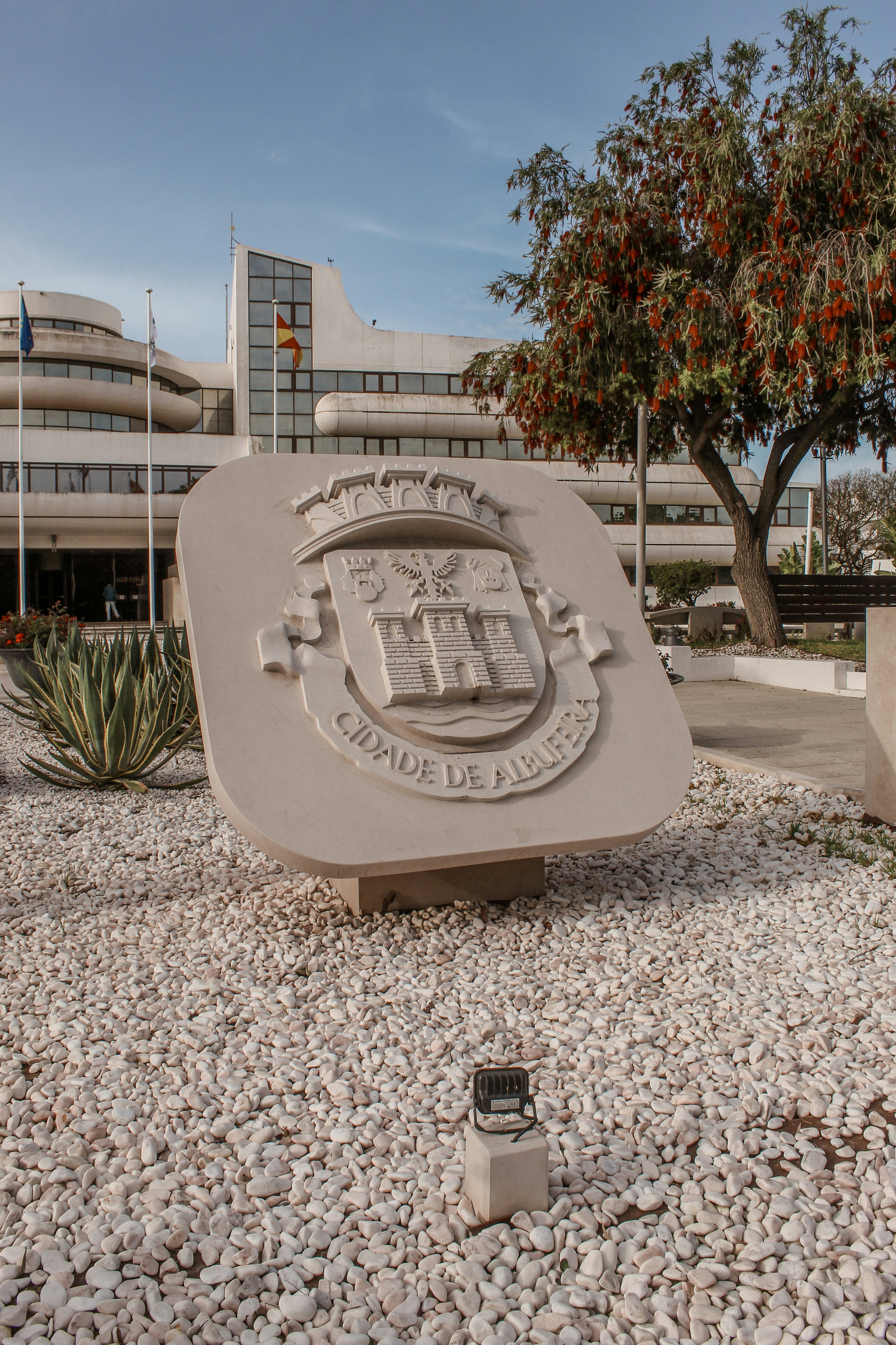 Leaned Monument: Crest carved in a sunlit plaza before a modern civic building