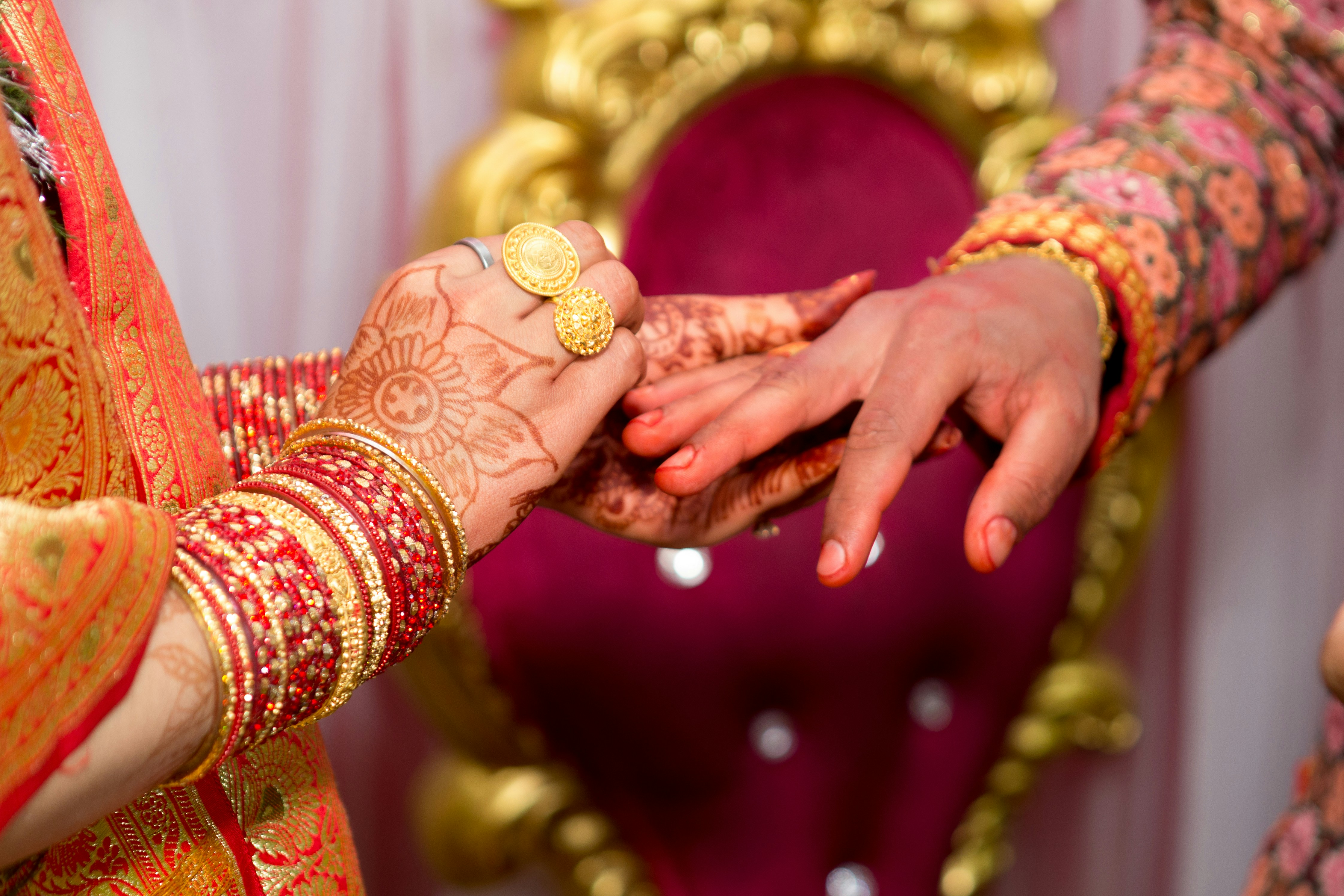 Close-up of two hands during a traditional wedding, adorned with ornate jewelry and henna, with a richly decorated red and gold attire and a purple throne in the background.