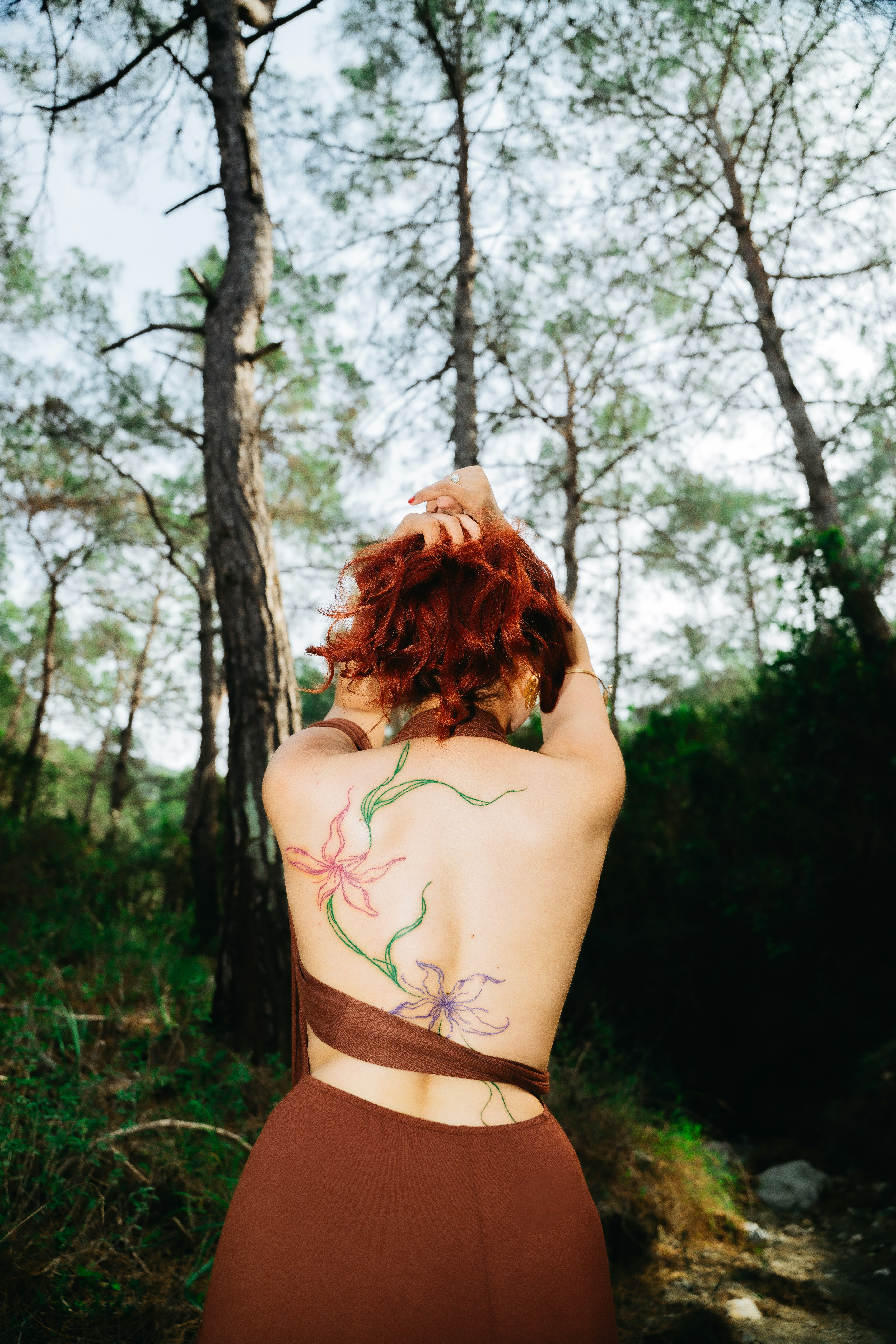 Red-haired figure in a sunlit pine forest, revealing a floral back tattoo