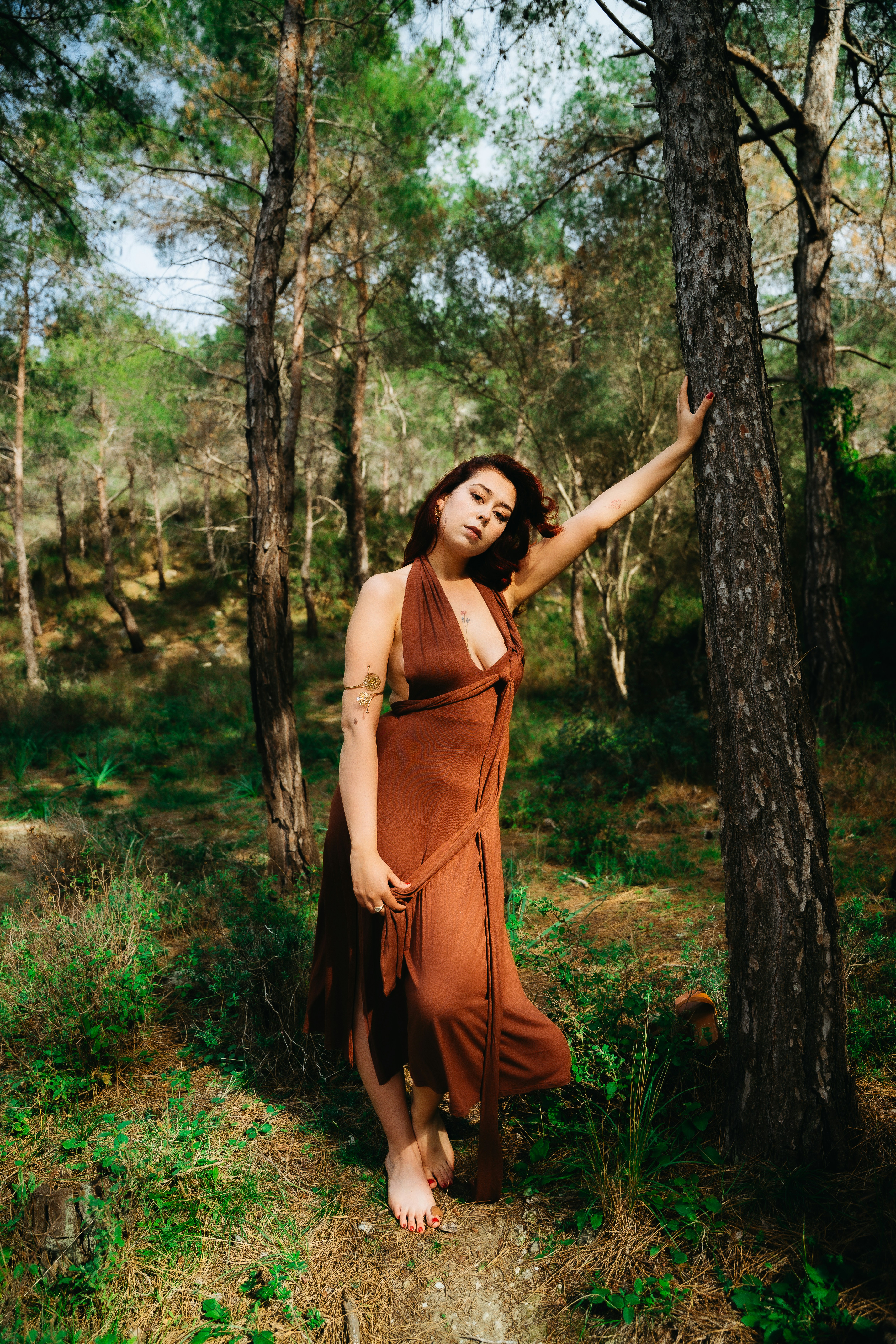 Brown dress, green forest: a woman leans against a pine tree in warm sunlight