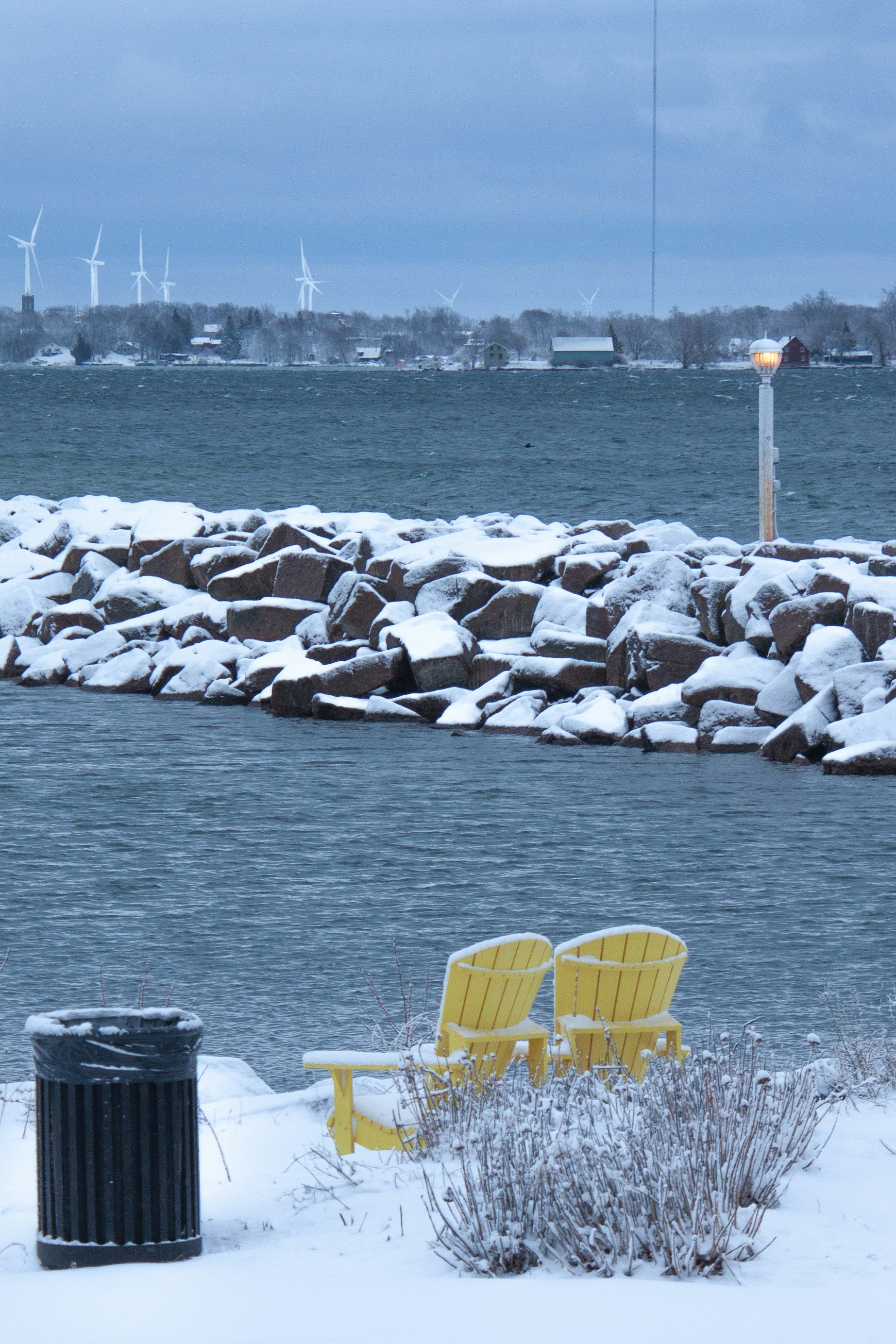 Snowy shoreline with yellow chairs facing a calm harbor