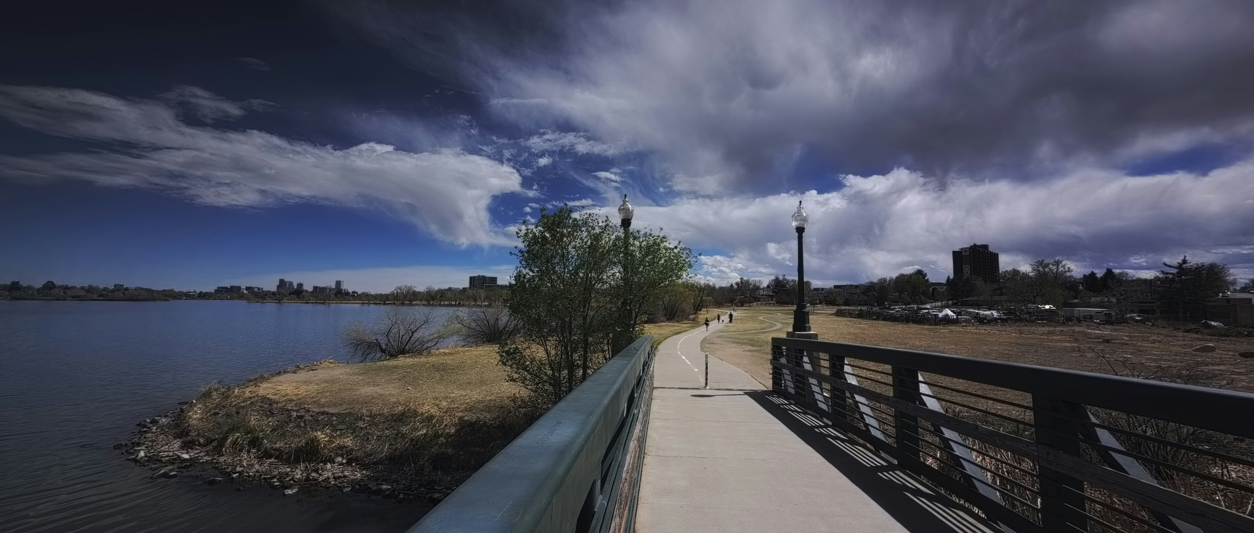 Pedestrian bridge over a calm river with a long railing, distant trees, and a dramatic sky; urban park scene captured in wide angle.