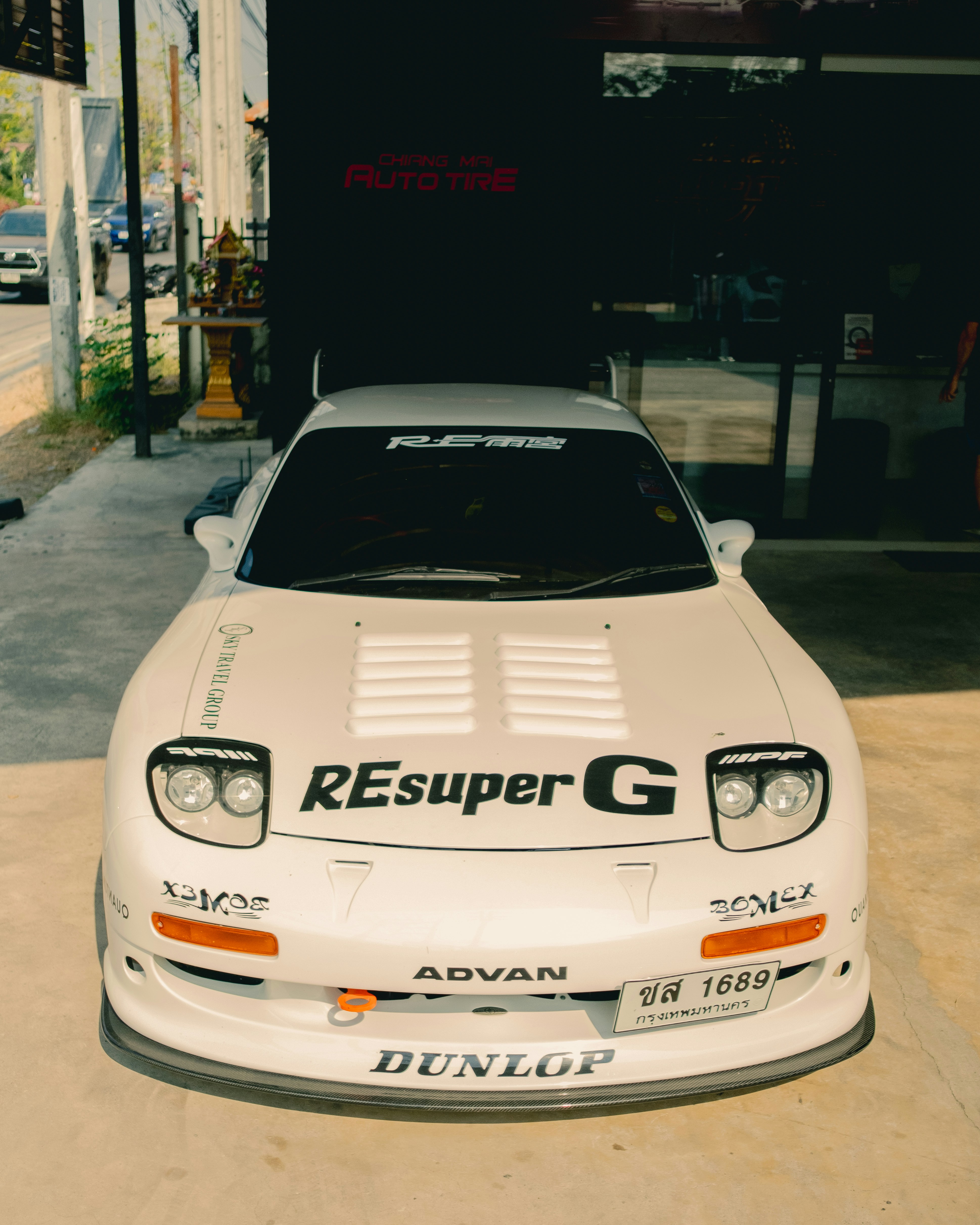A white racecar with bold branding sits at a shop entrance, the front bumper and hood featuring sponsor logos and a large 'REsuper G' decal under the windshield. The scene is captured in daylight with the car centered in the frame.