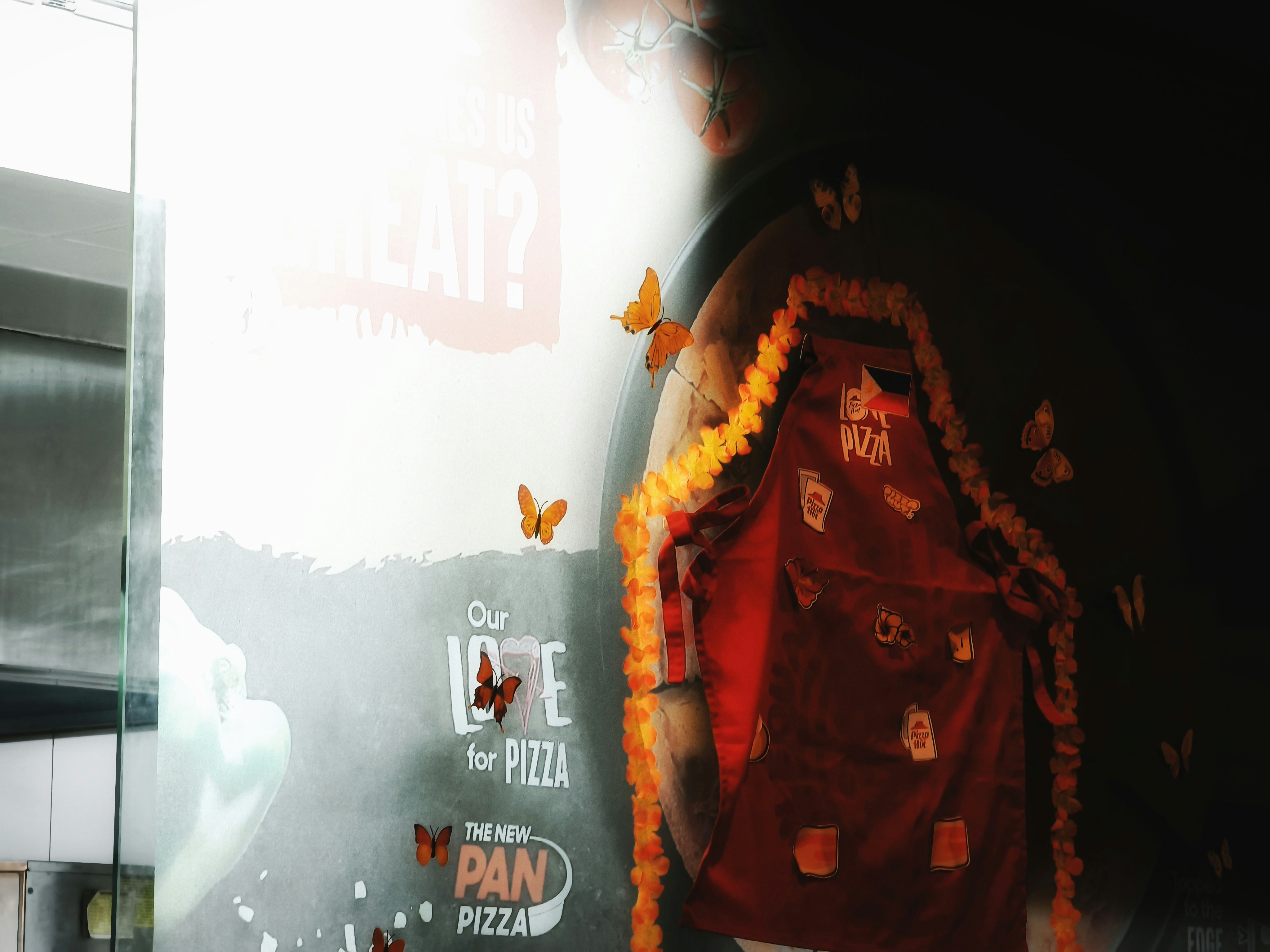 A storefront mural featuring a pizza shrine framed by bright butterflies and orange garlands, with soft backlighting and a graffiti-like backdrop.