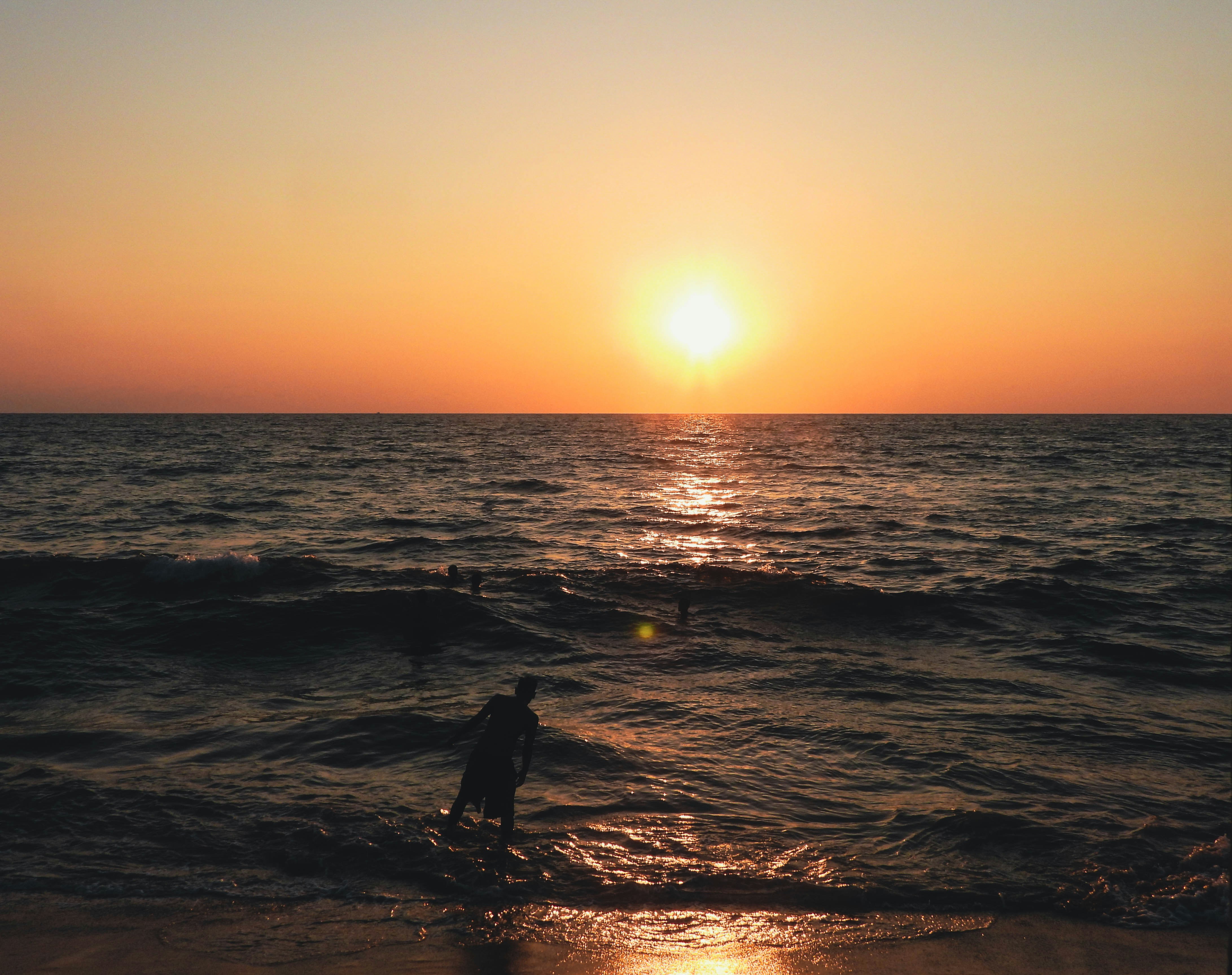 Silhouette of a person with a surfboard near the shoreline during a warm sunset over a calm sea; low-light scene with a vivid gradient sky and reflective water surface.