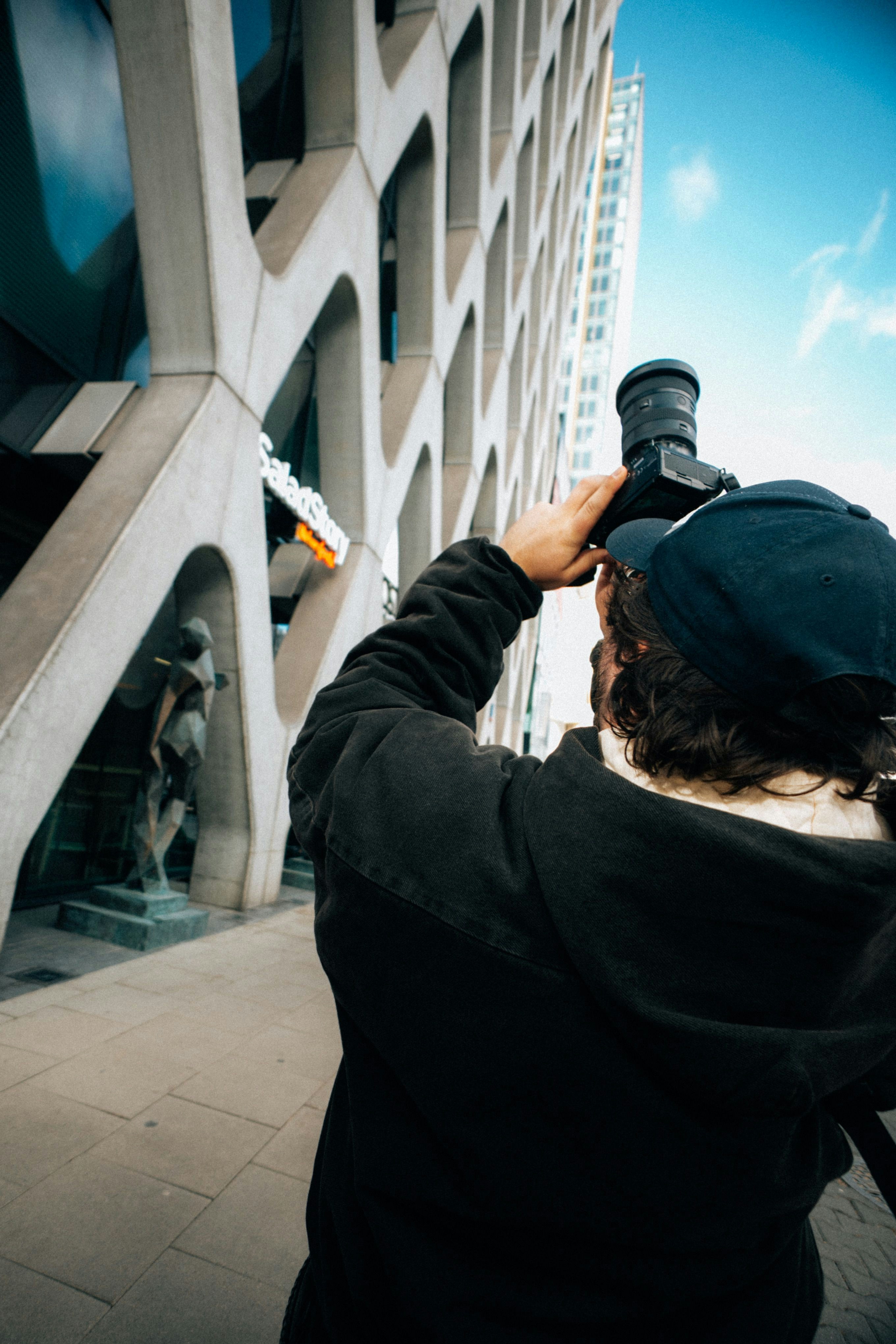 Camera Cradle: Street Photographer at the Corner of Concrete and Sky