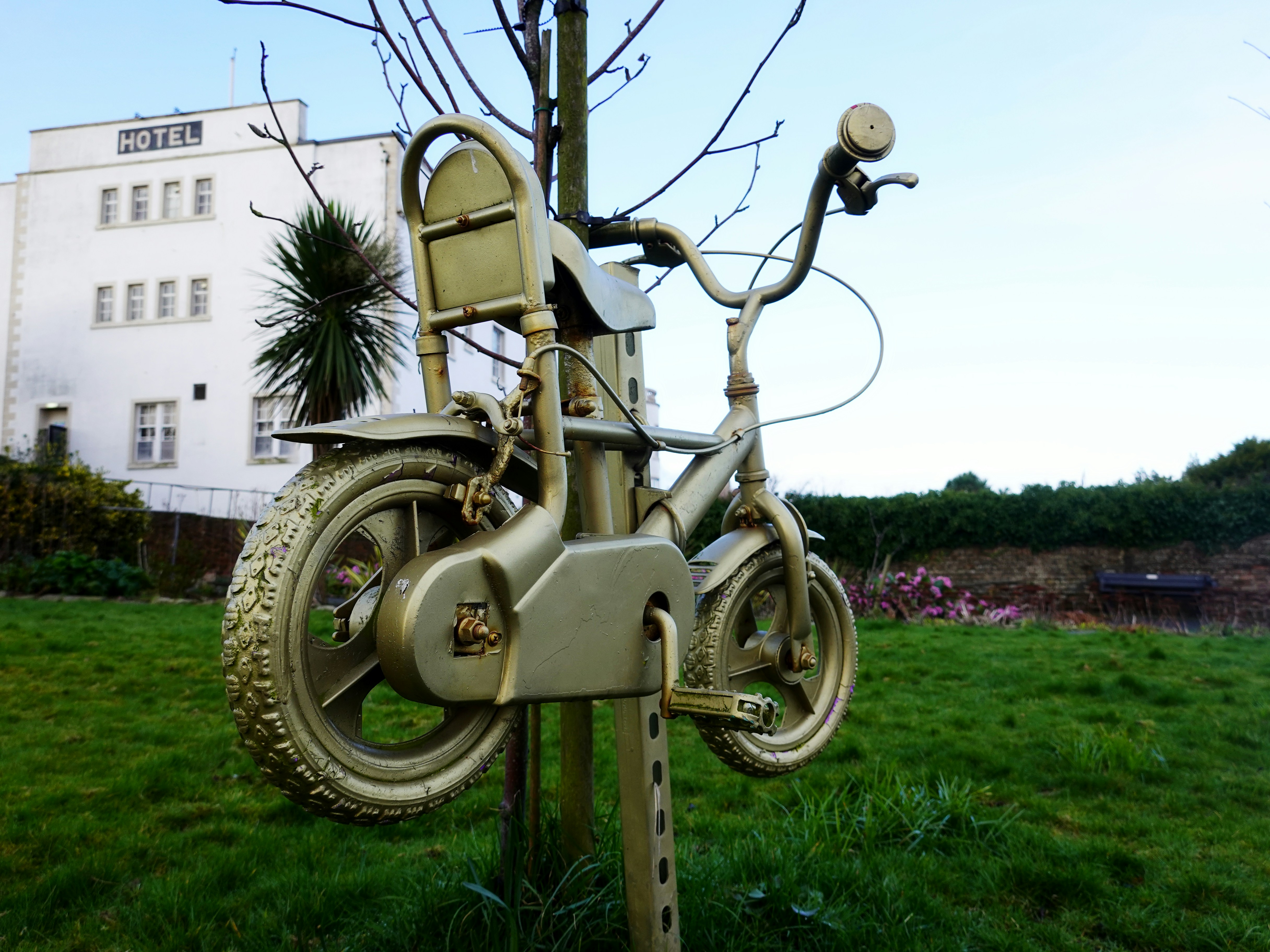 Deserted moped sculpture anchored in a park with a distant hotel façade
