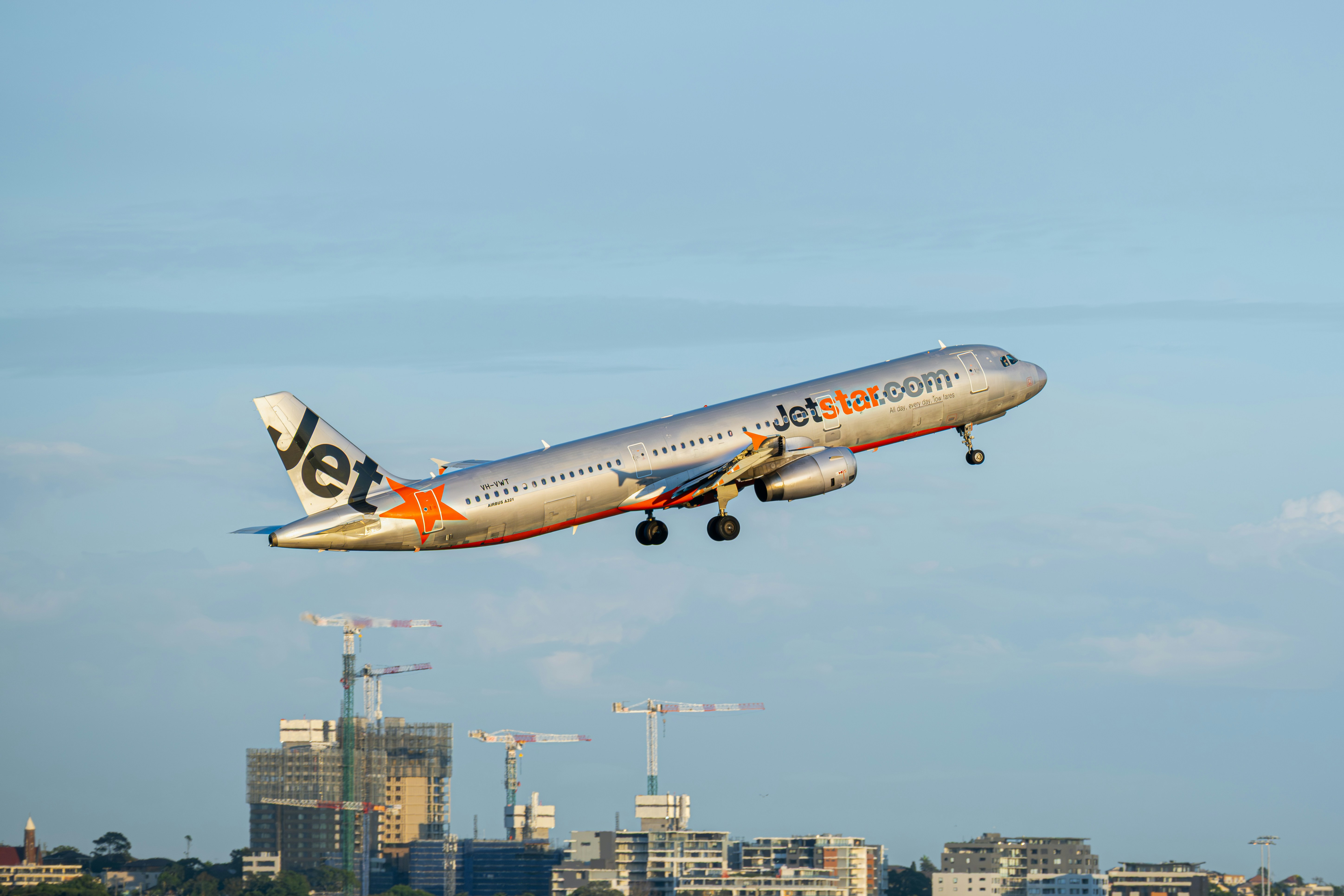 A Airbus A321 climbs after takeoff, with its landing gear still extended. The aircraft is seen against a clear sky, with cranes and buildings visible below in the background.