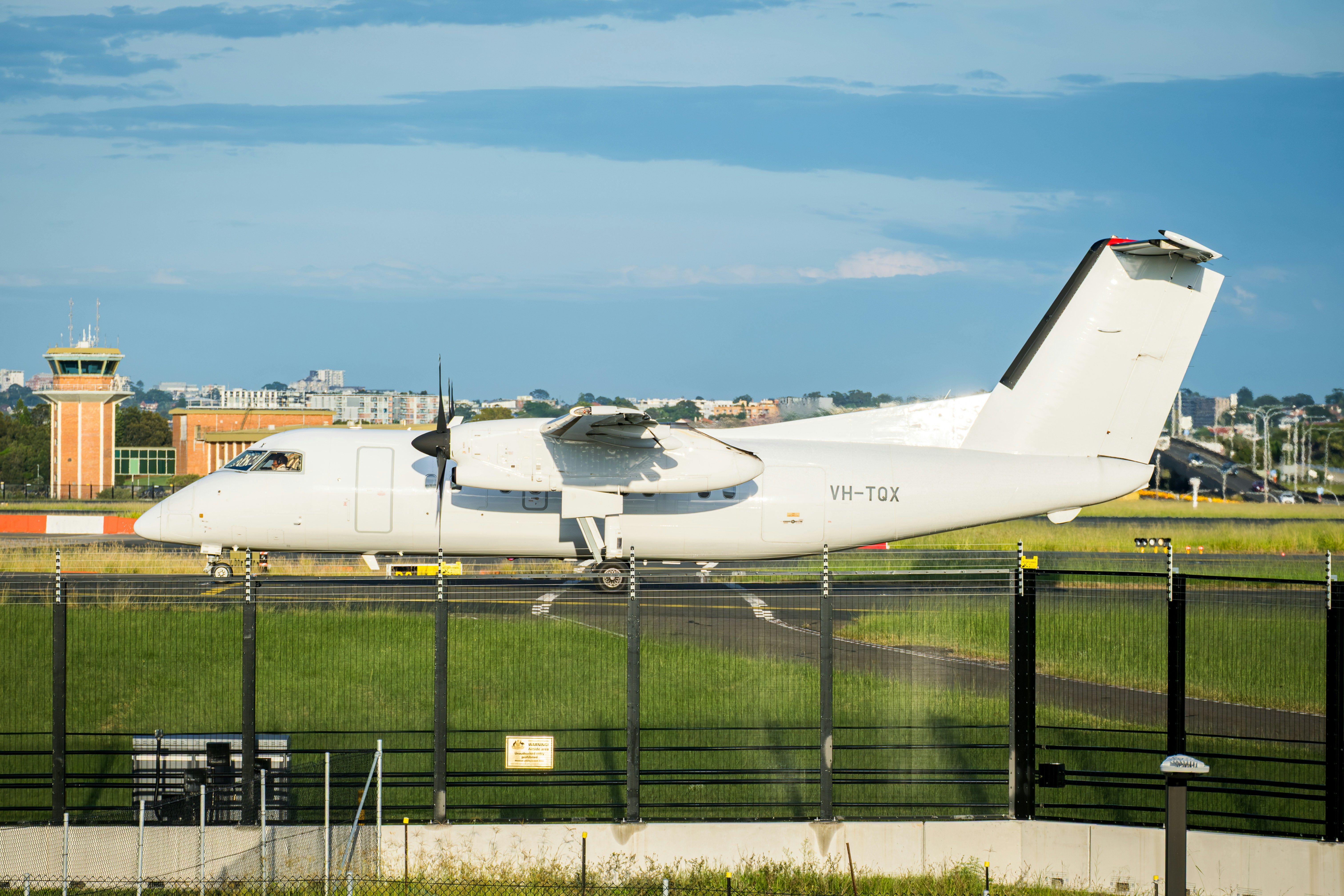 White twin-engine turboprop jet with tail number VH-TOX sits behind a fence at an airfield