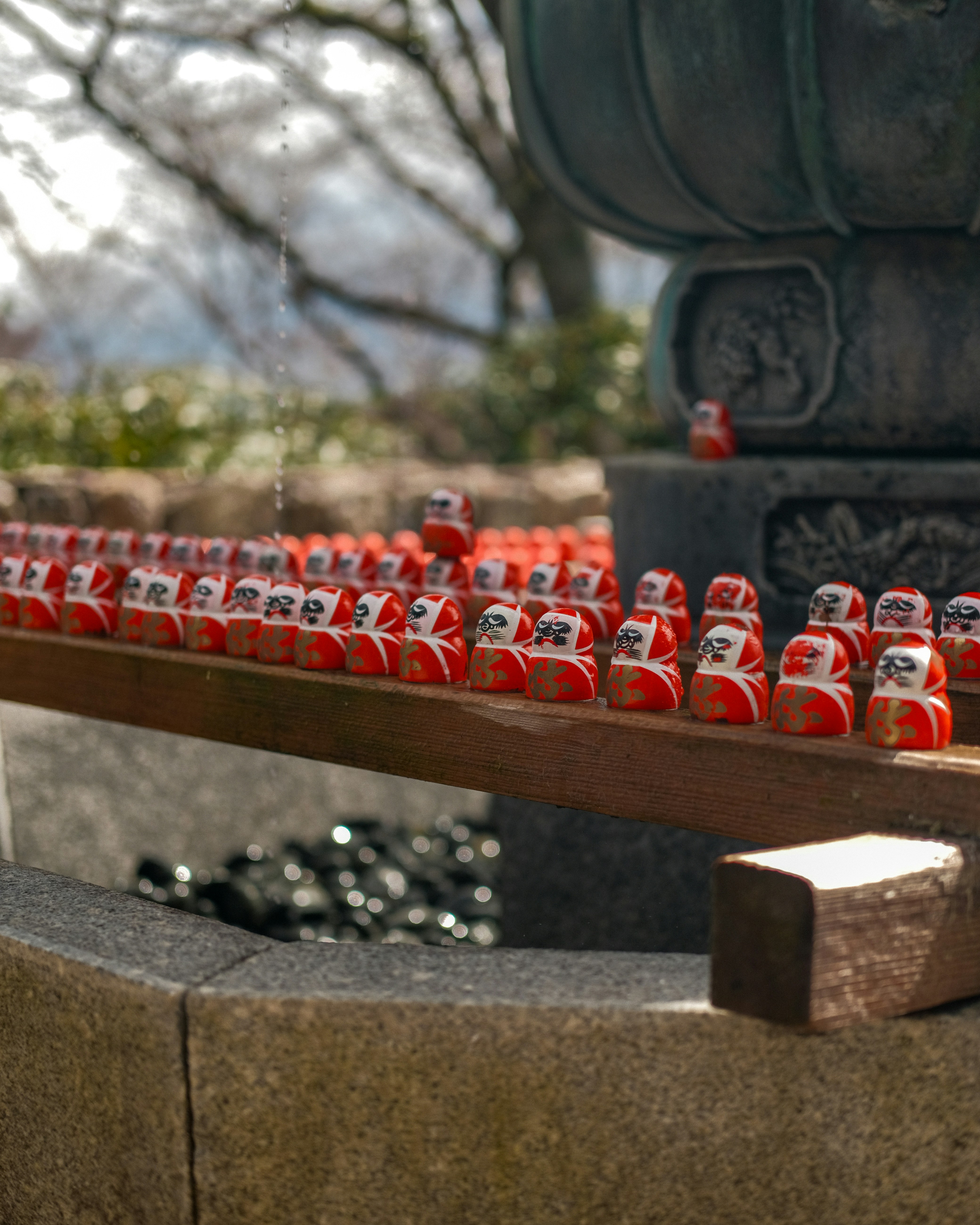 Row of red and white daruma dolls lining a wooden railing at a stone basin