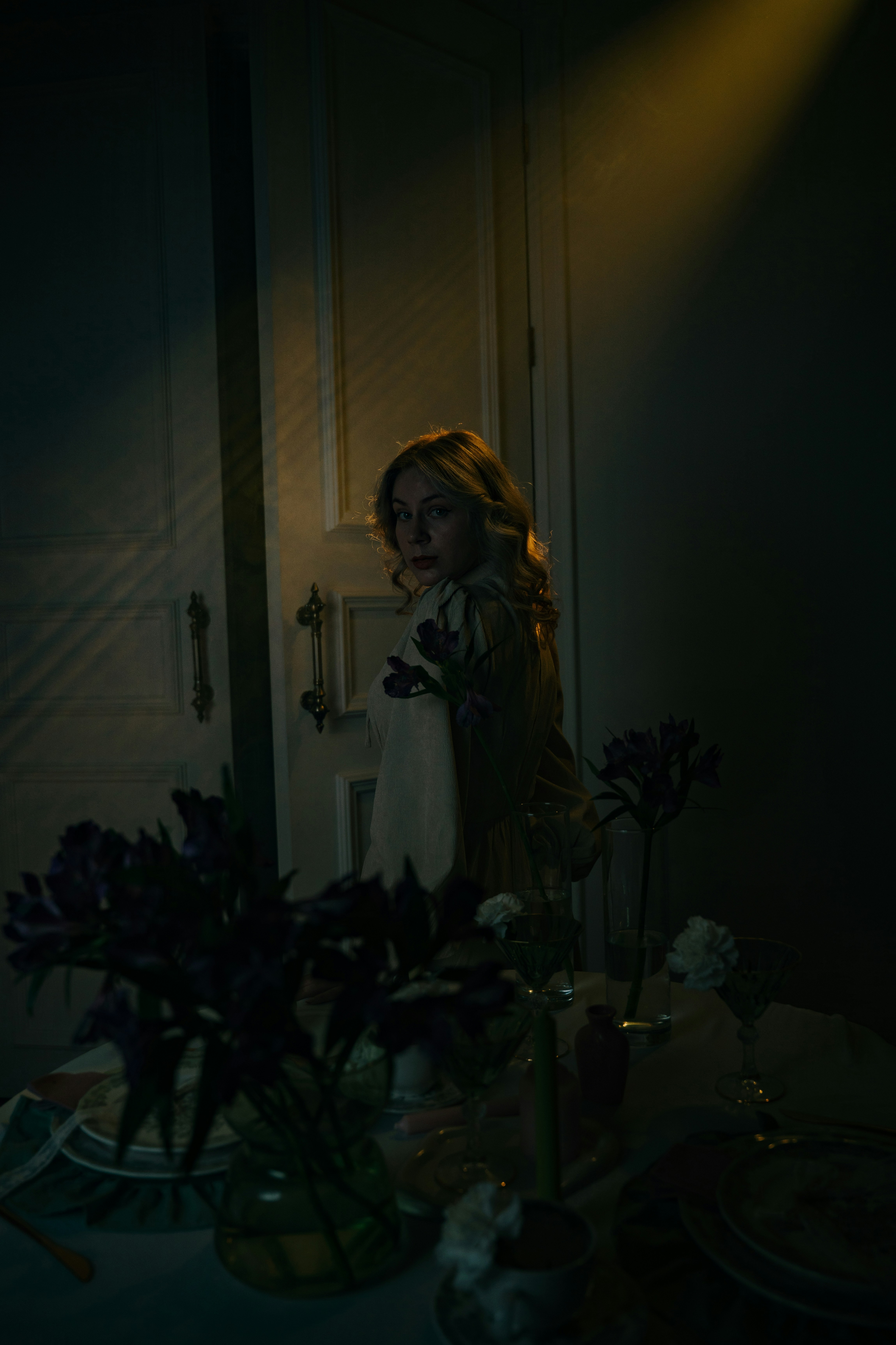 Dimly lit intimate scene featuring a woman standing by a doorway at the end of a table set for a meal; a solitary spotlight highlights her face and upper body, with floral arrangements and dinnerware adding context. The composition emphasizes mood and atmosphere rather than action.
