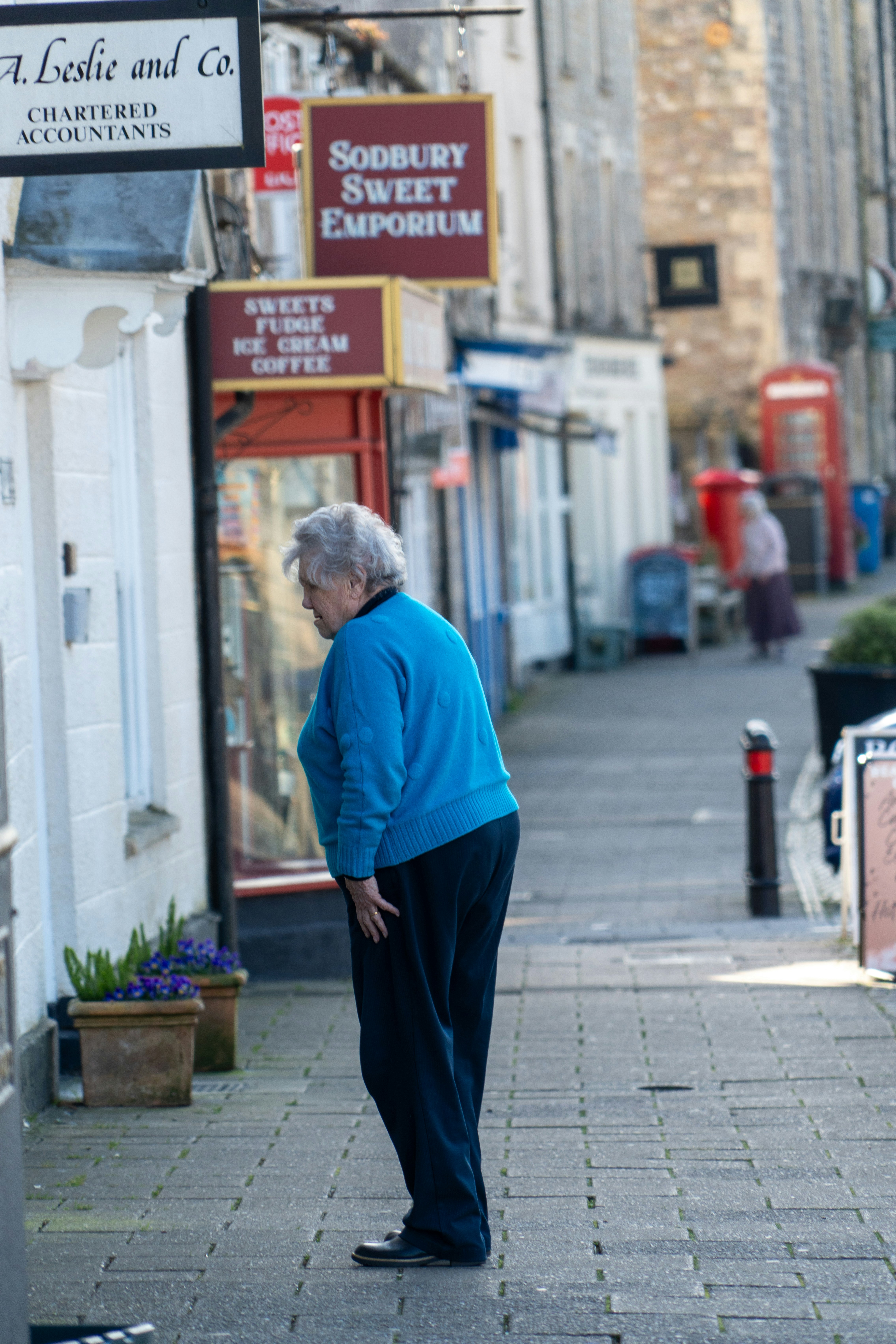 Older woman in a bright blue sweater bends slightly as she surveys a shopfront on a busy pedestrian street lined with storefront signs and a flower pot in the foreground. A soft, shallow depth of field emphasizes her presence against the urban backdrop.