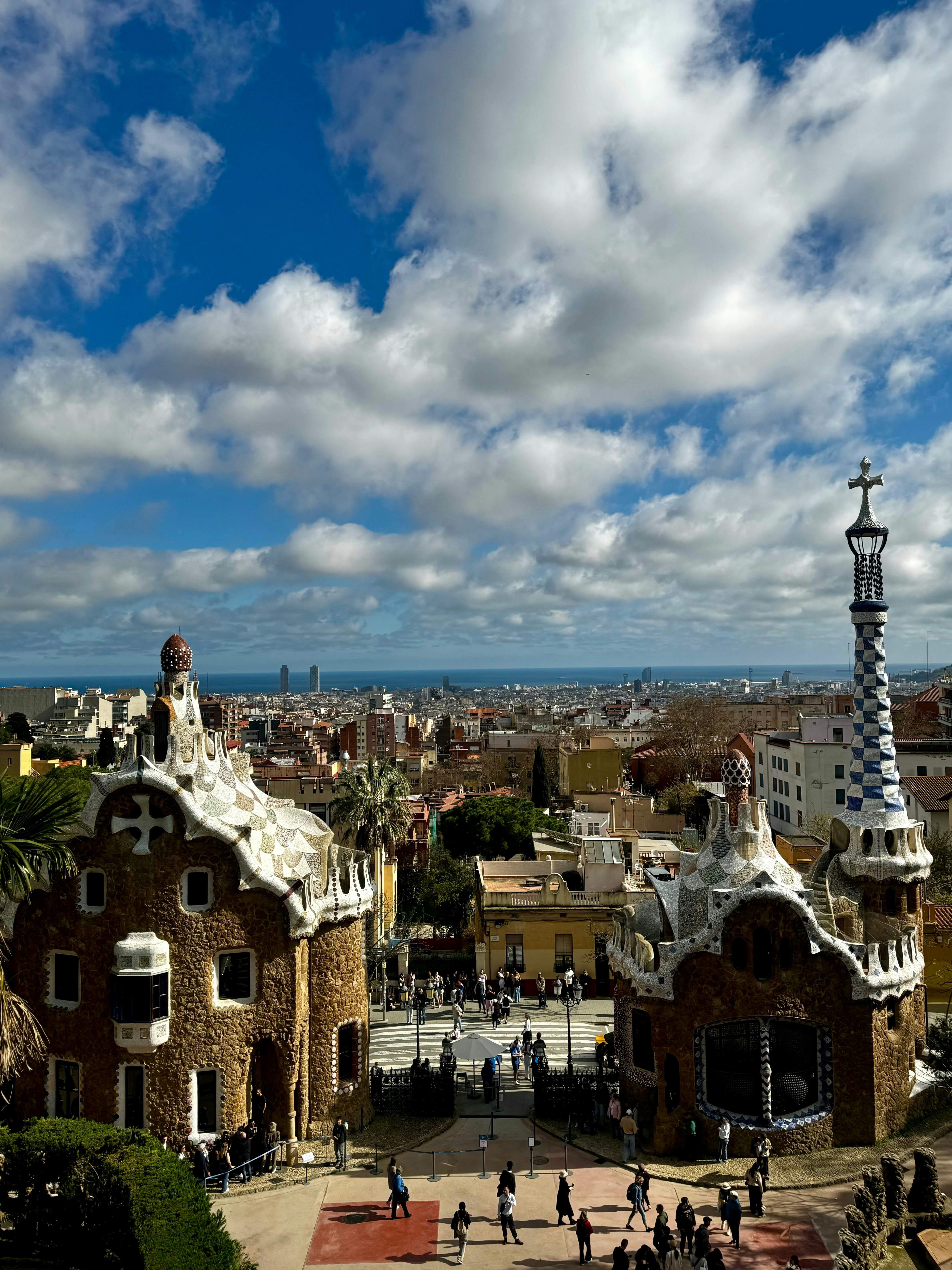 Colorful architectural detail at Parc Güell with undulating rooftops and a stairway crowd, overlooking a cityscape toward the sea.