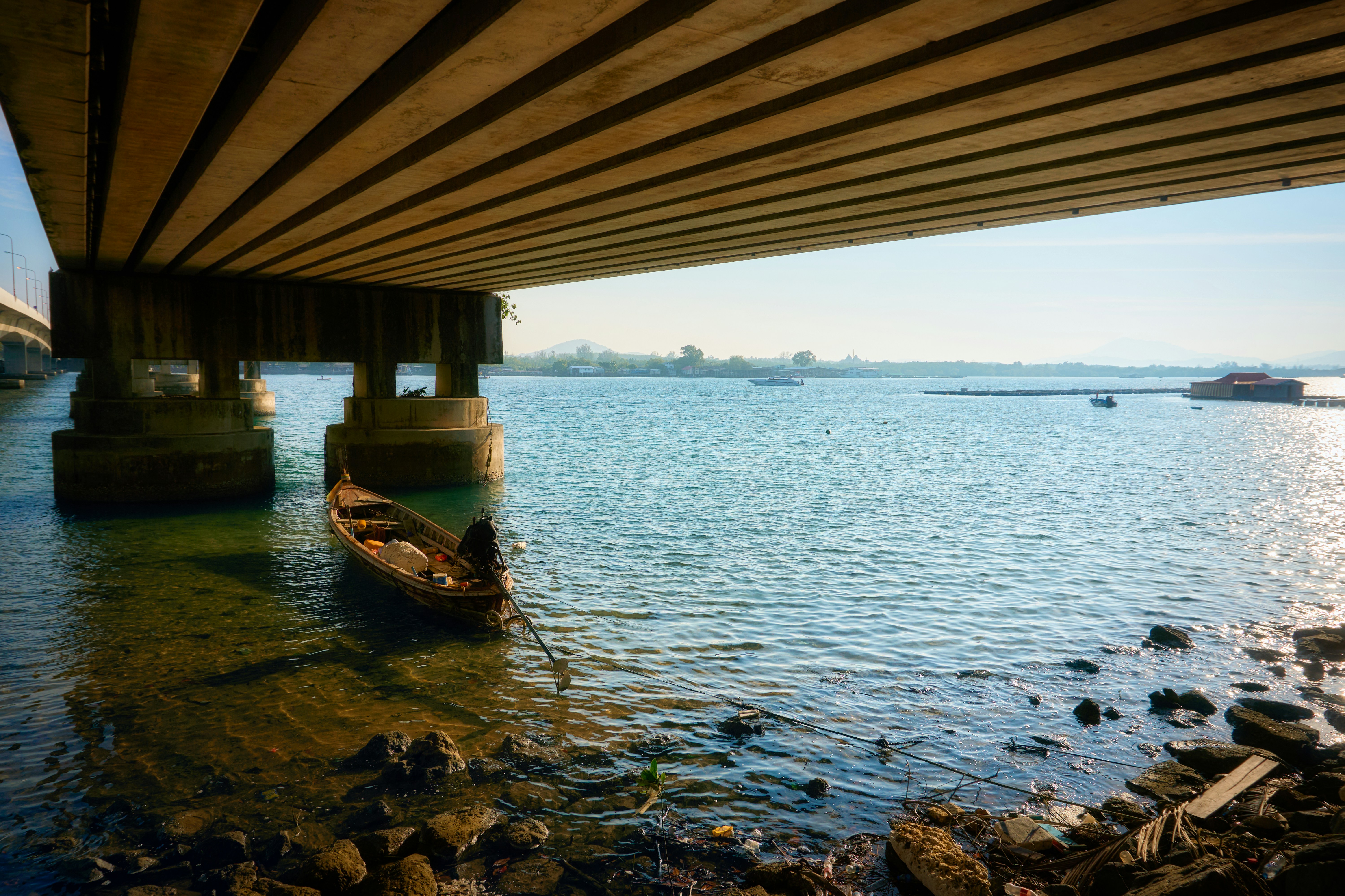 Boat resting under a sunlit bridge, meeting the river at rocky shore