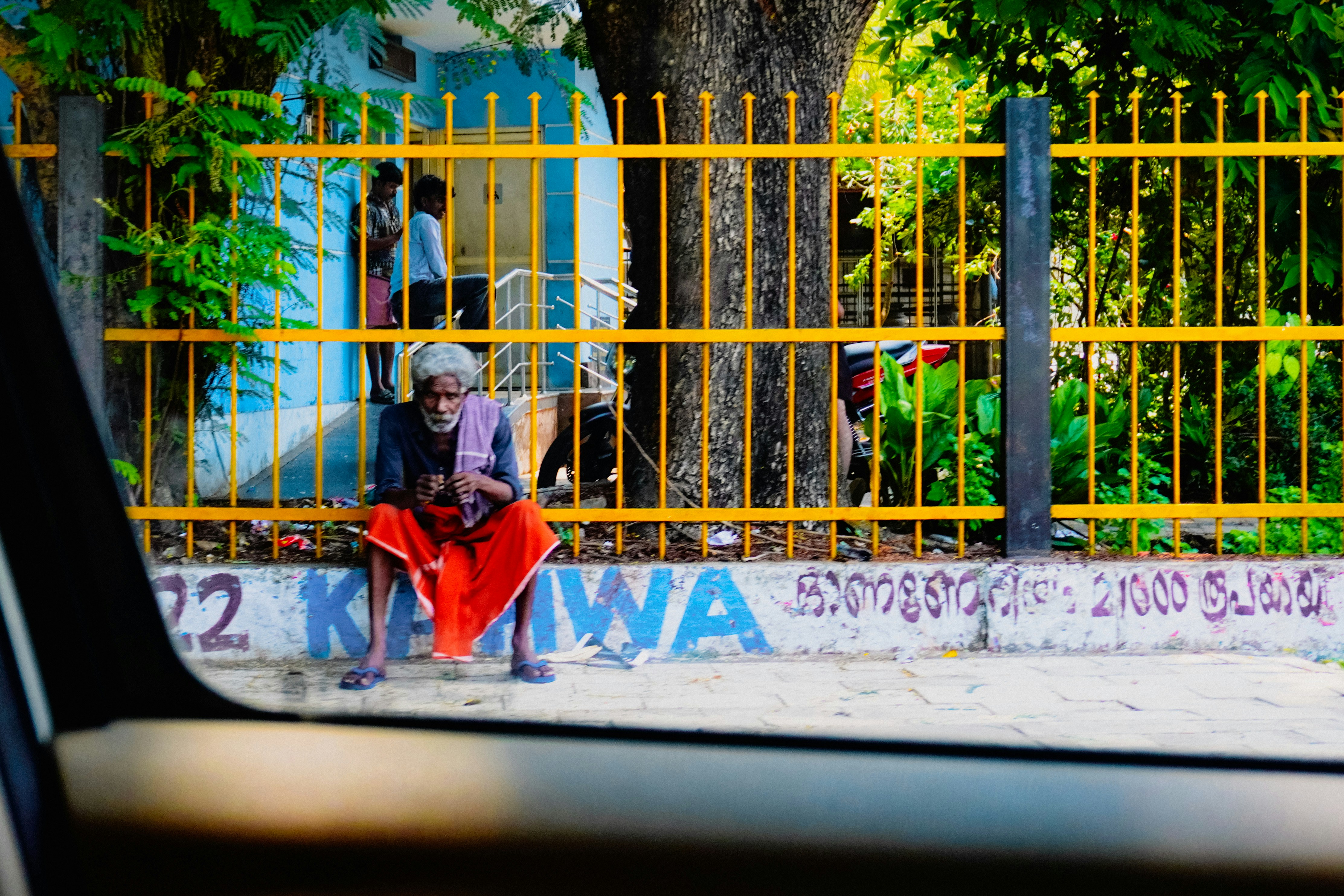Man in bright red sarong sits by a yellow fence watching the street