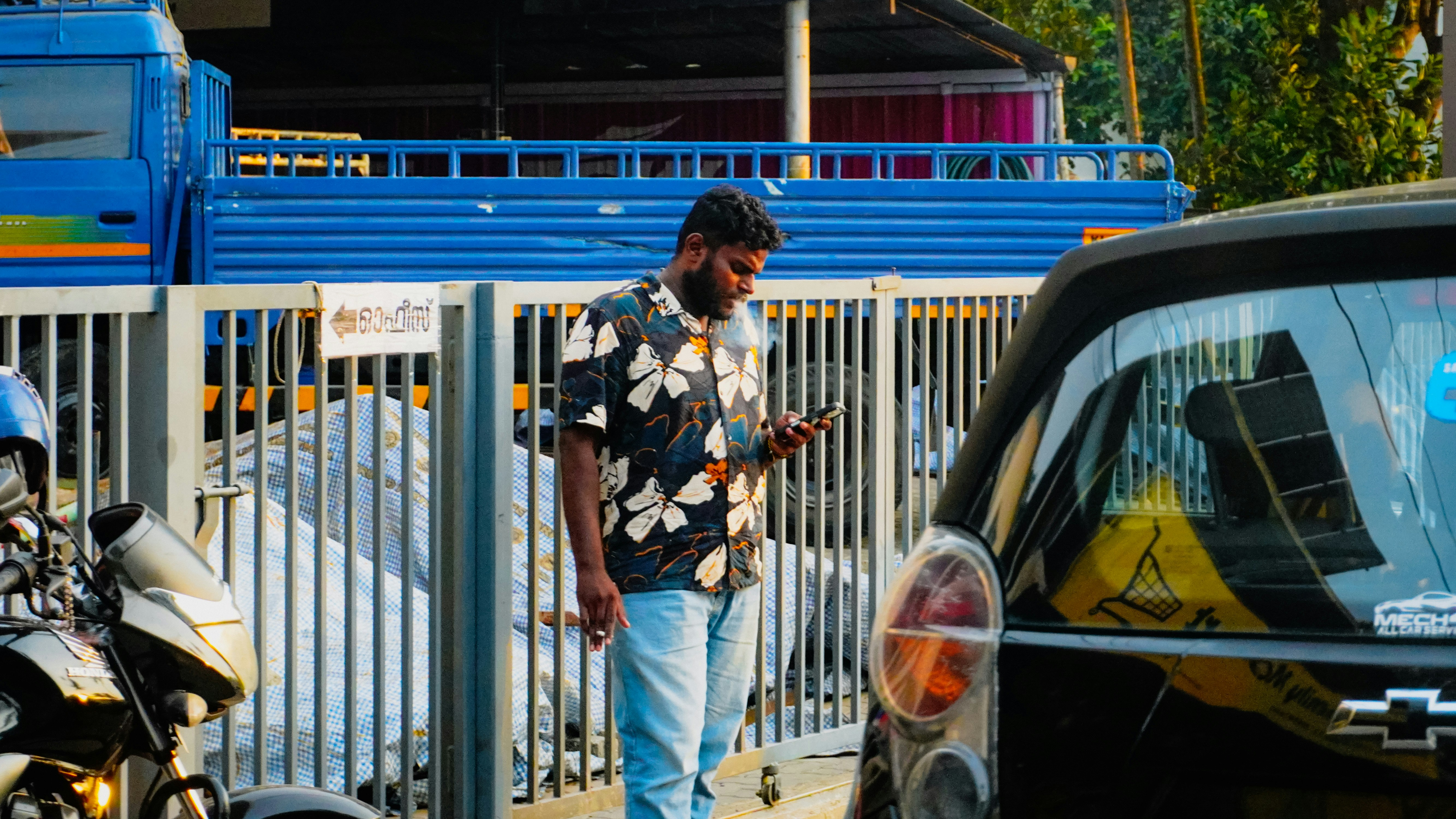 Man with Floral Shirt Stands by a Bus Yard, Glancing at His Phone