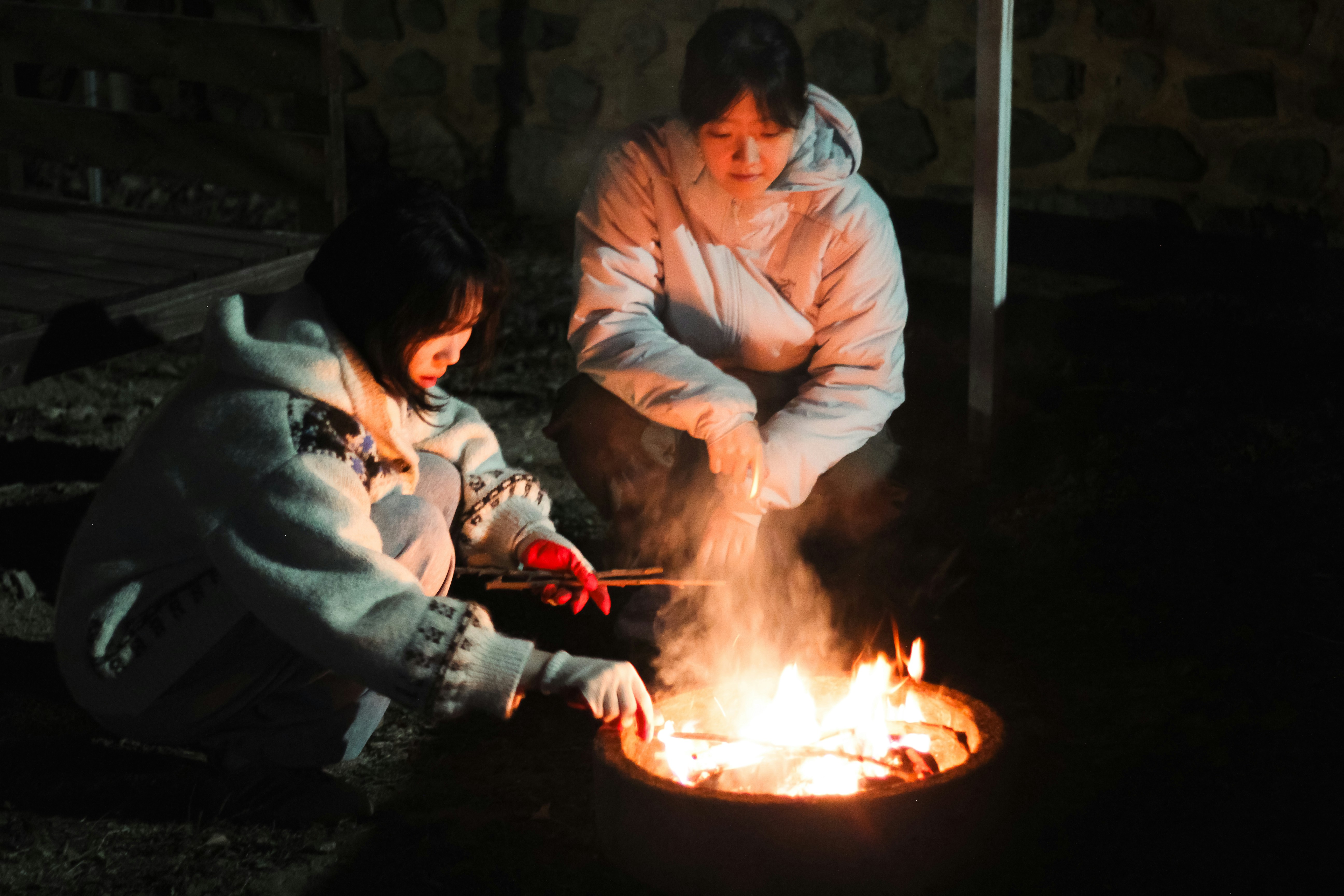 Two silhouettes tending a crackling fire under a dim night sky