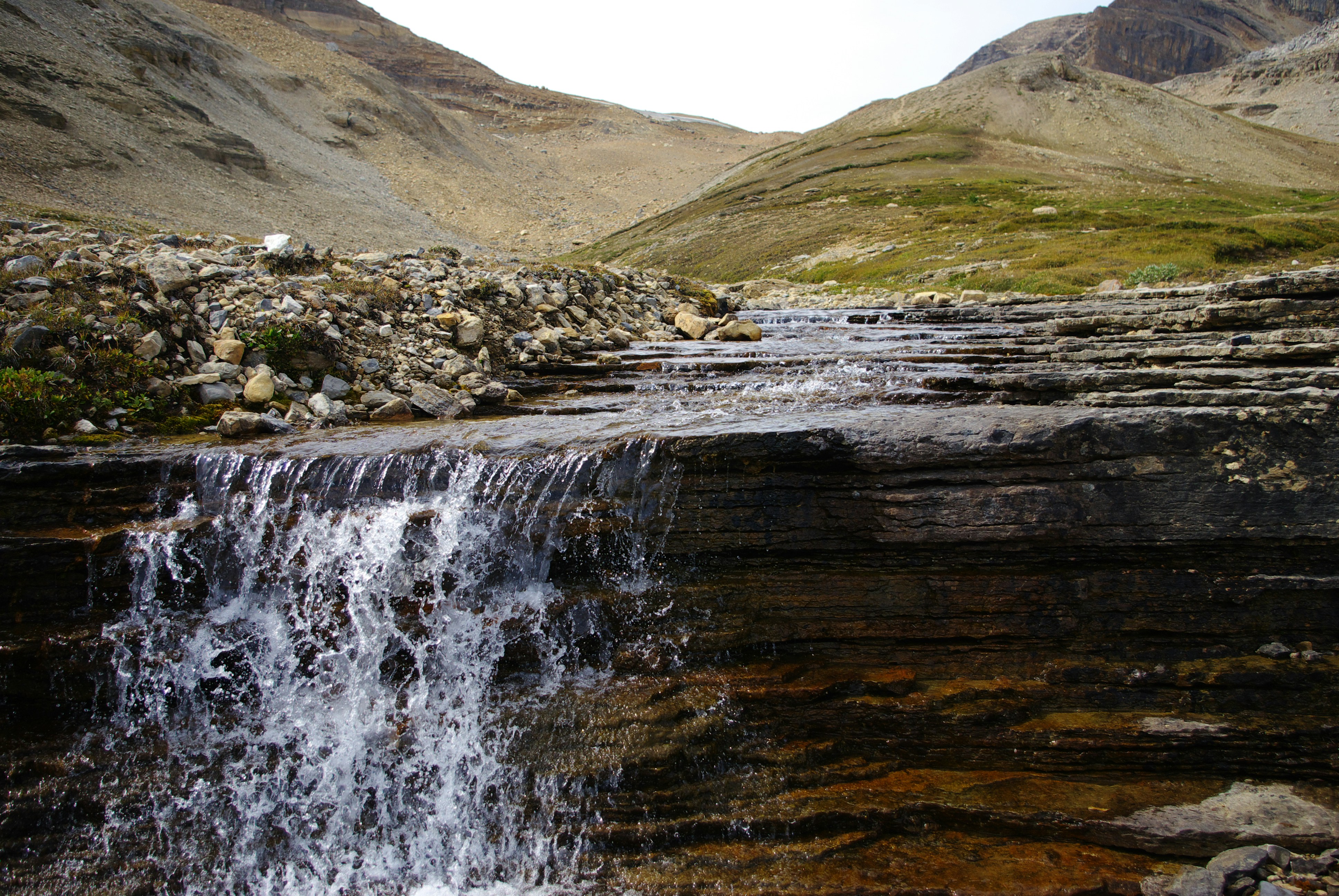 Water spills over layered rock at the edge of a rocky alpine stream, with a distant slope and sparse vegetation in a muted, grey-brown palette.