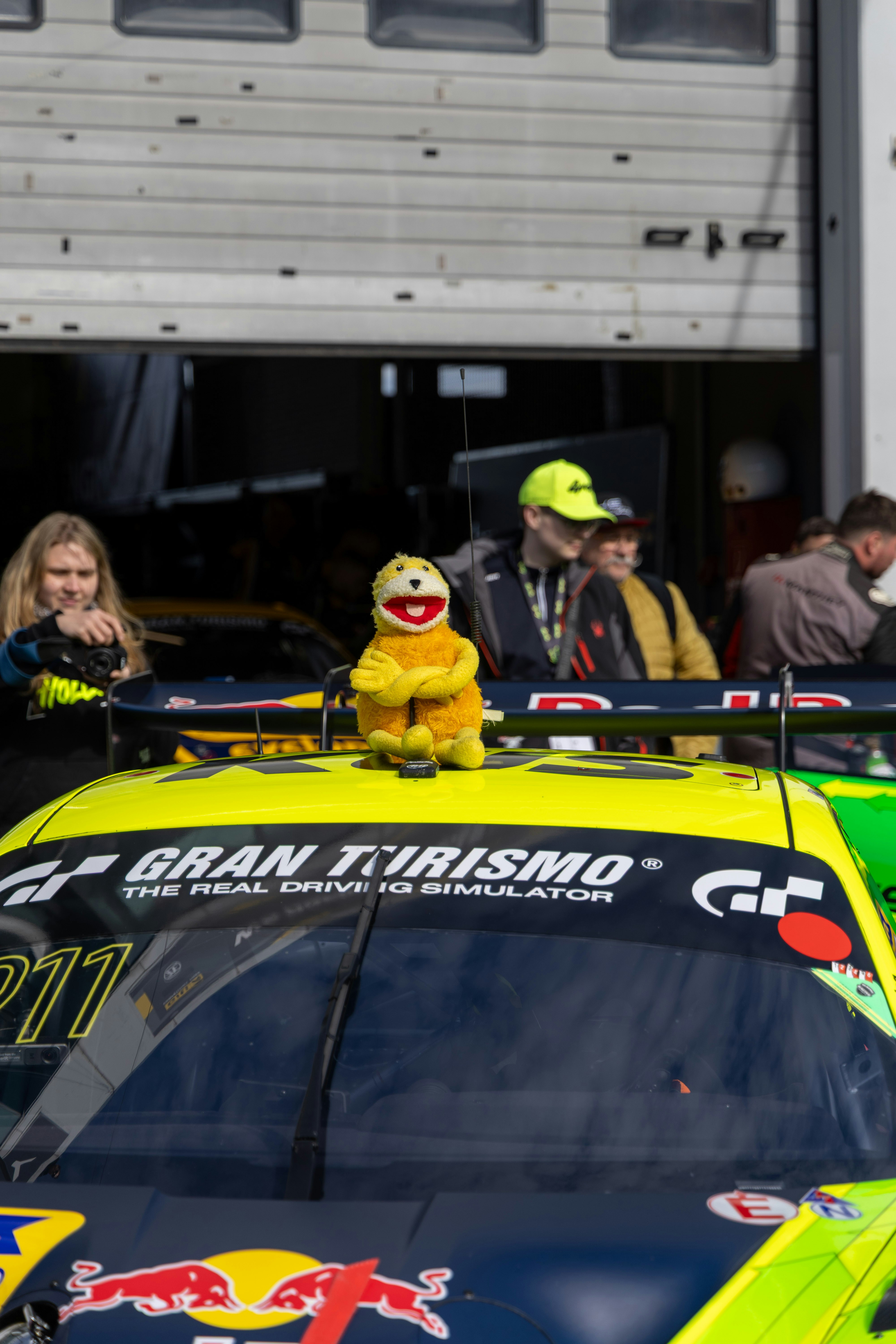 Bright yellow stuffed muppet sits atop a neon race car, with pit crew and spectators blurred in the background during a race day event.