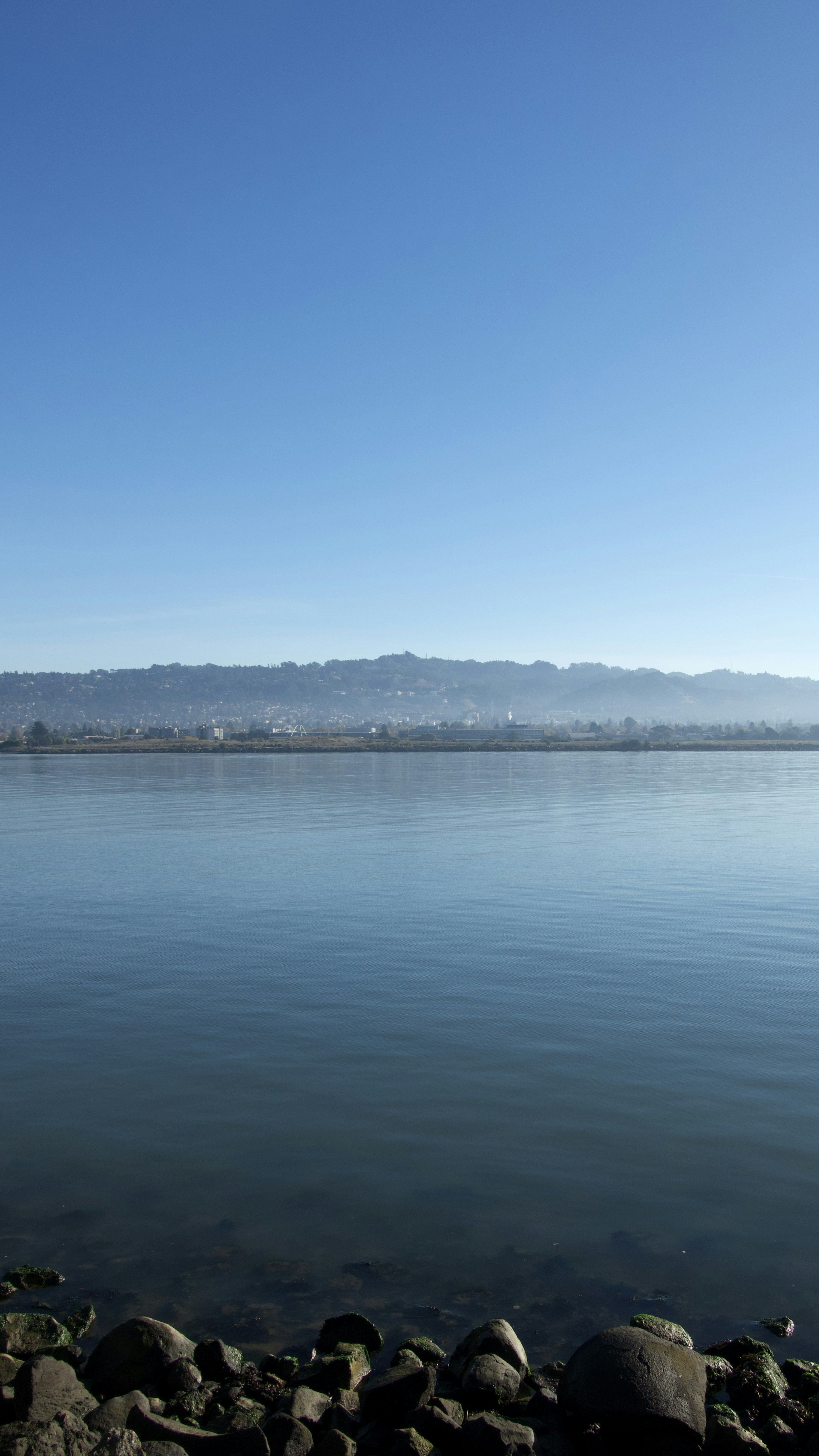 Calm waters mirror a clear blue sky with a distant town and hills on the horizon; rocky shore anchors the foreground. This landscape emphasizes tranquil mood and wide-scale perspective.