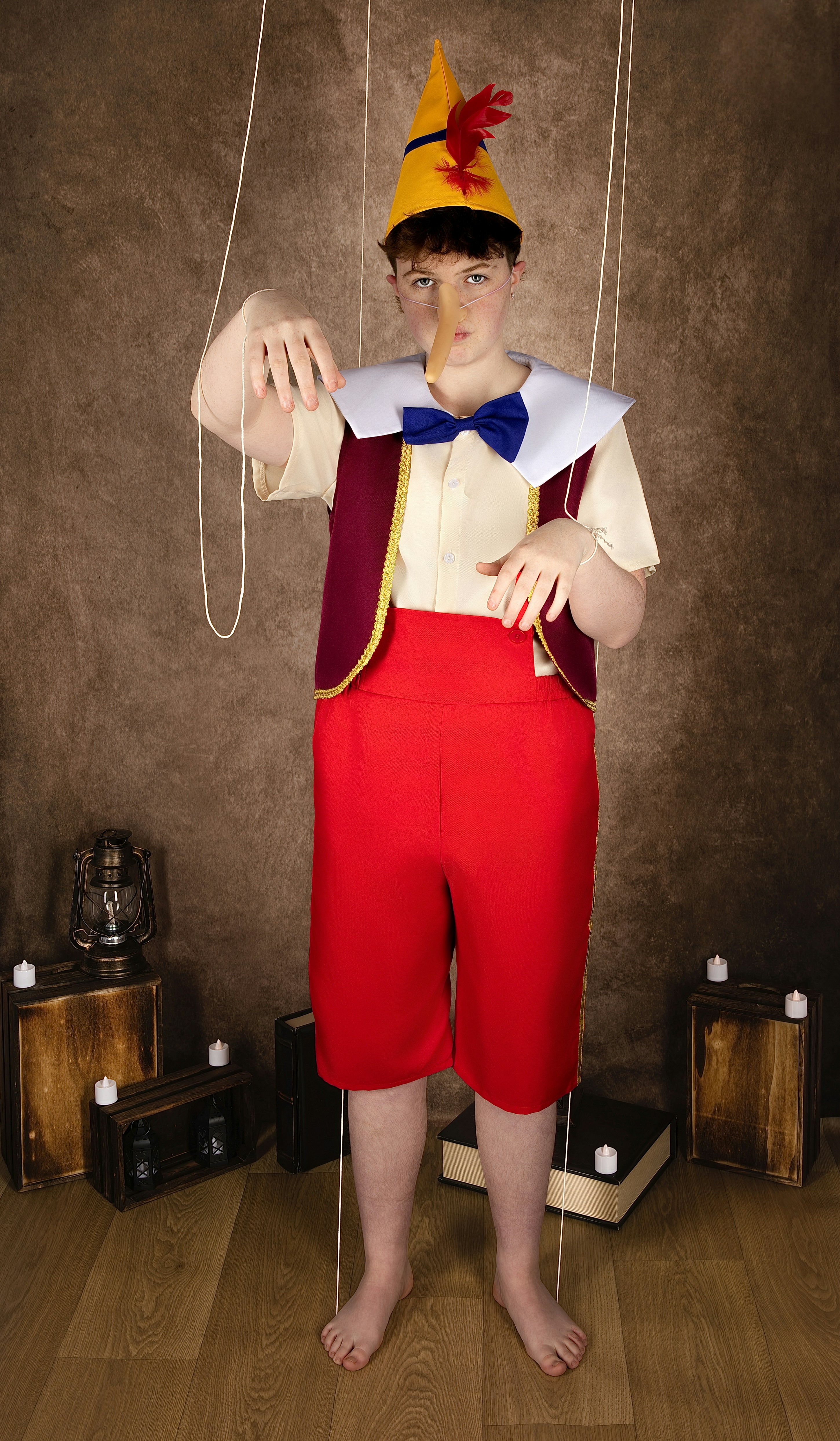 Young performer in a homemade costume stands barefoot among props, strings rises above as if puppeteered. A quirky scene with a party hat, bow tie, and red shorts against a brown textured backdrop.