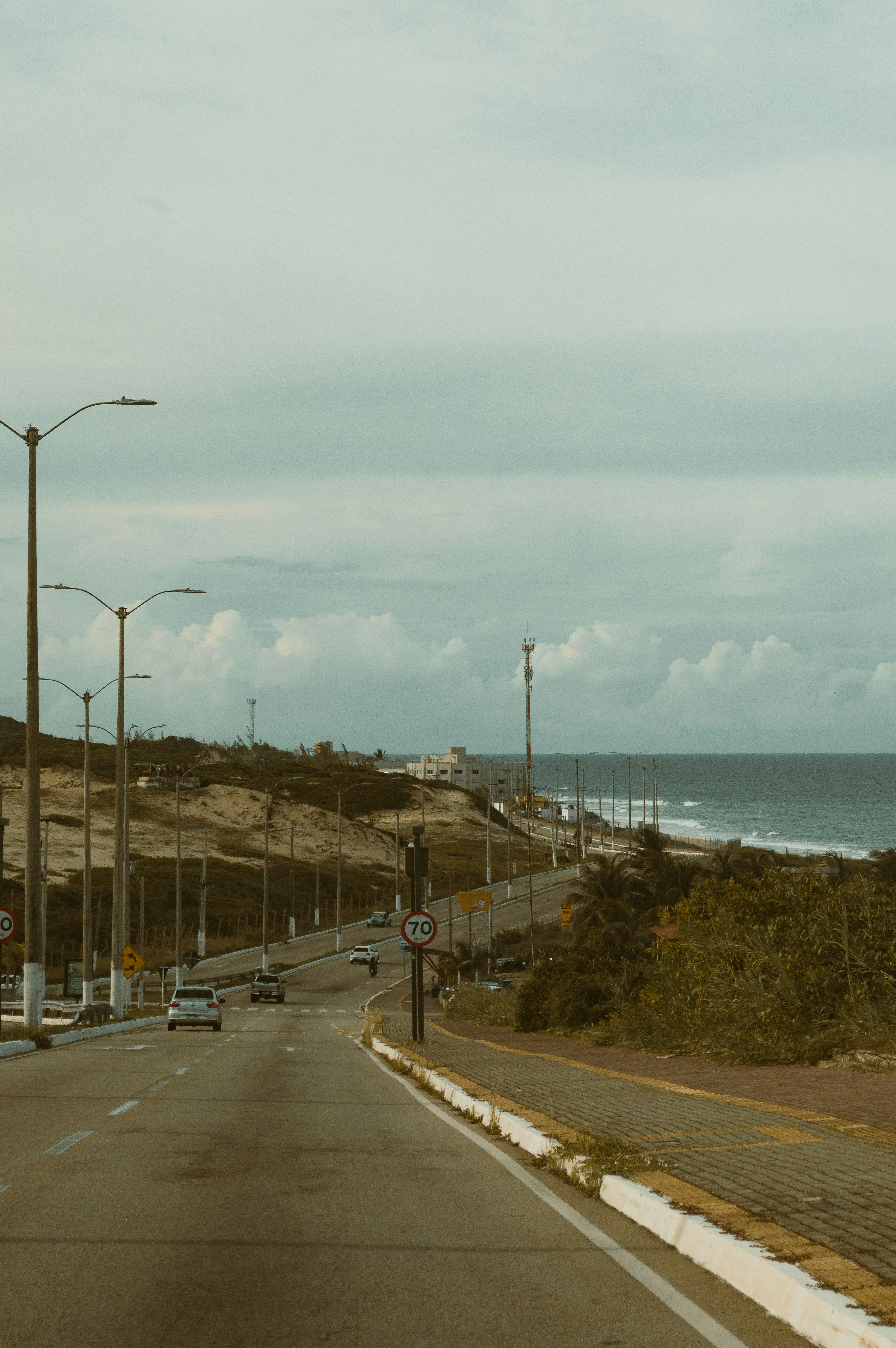 Long coastal road recedes toward the horizon with lamp posts and a calm sea on the right; subdued tones and soft light evoke a tranquil seascape.