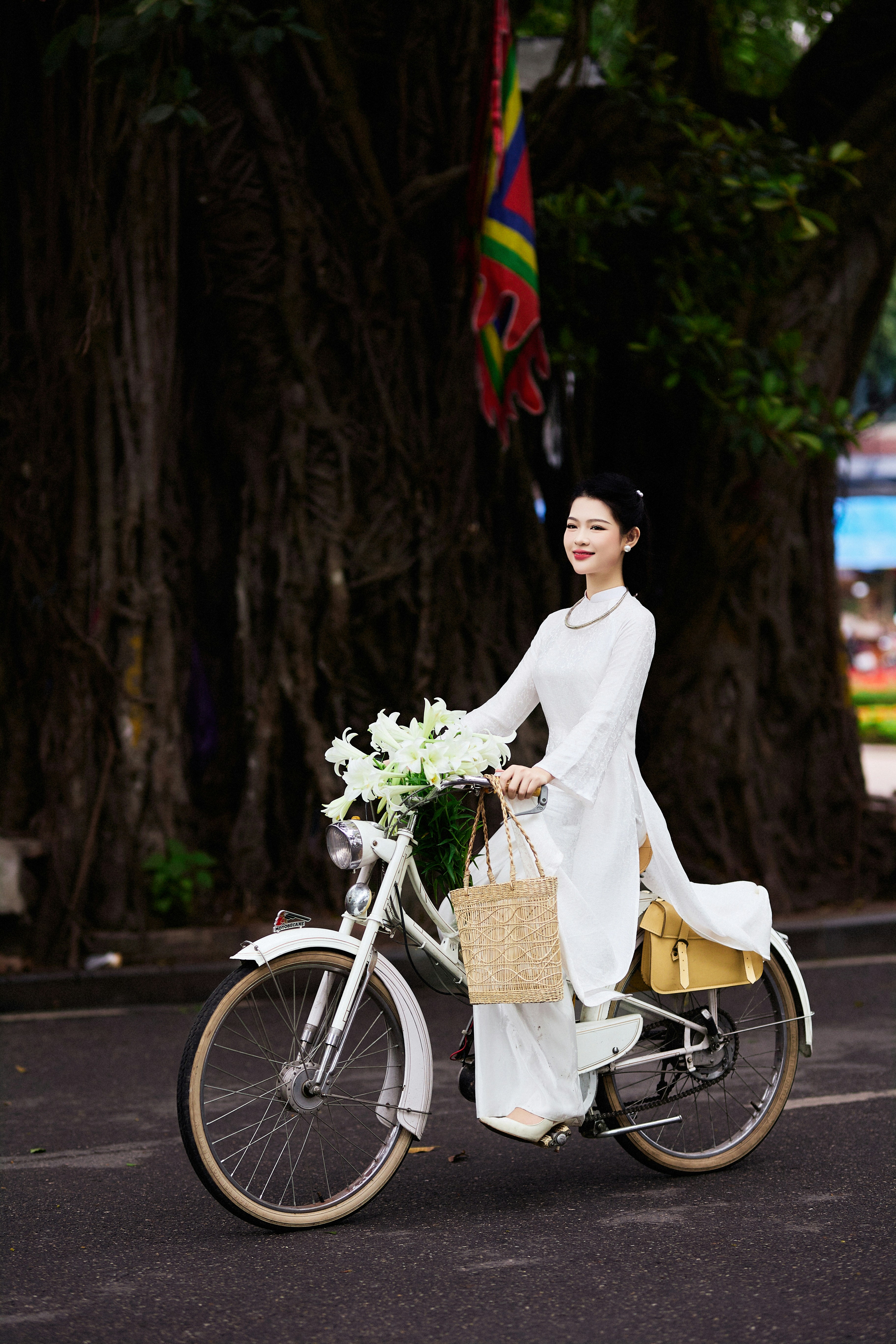 White-gowned rider with bouquet on a vintage bicycle beneath a towering tree