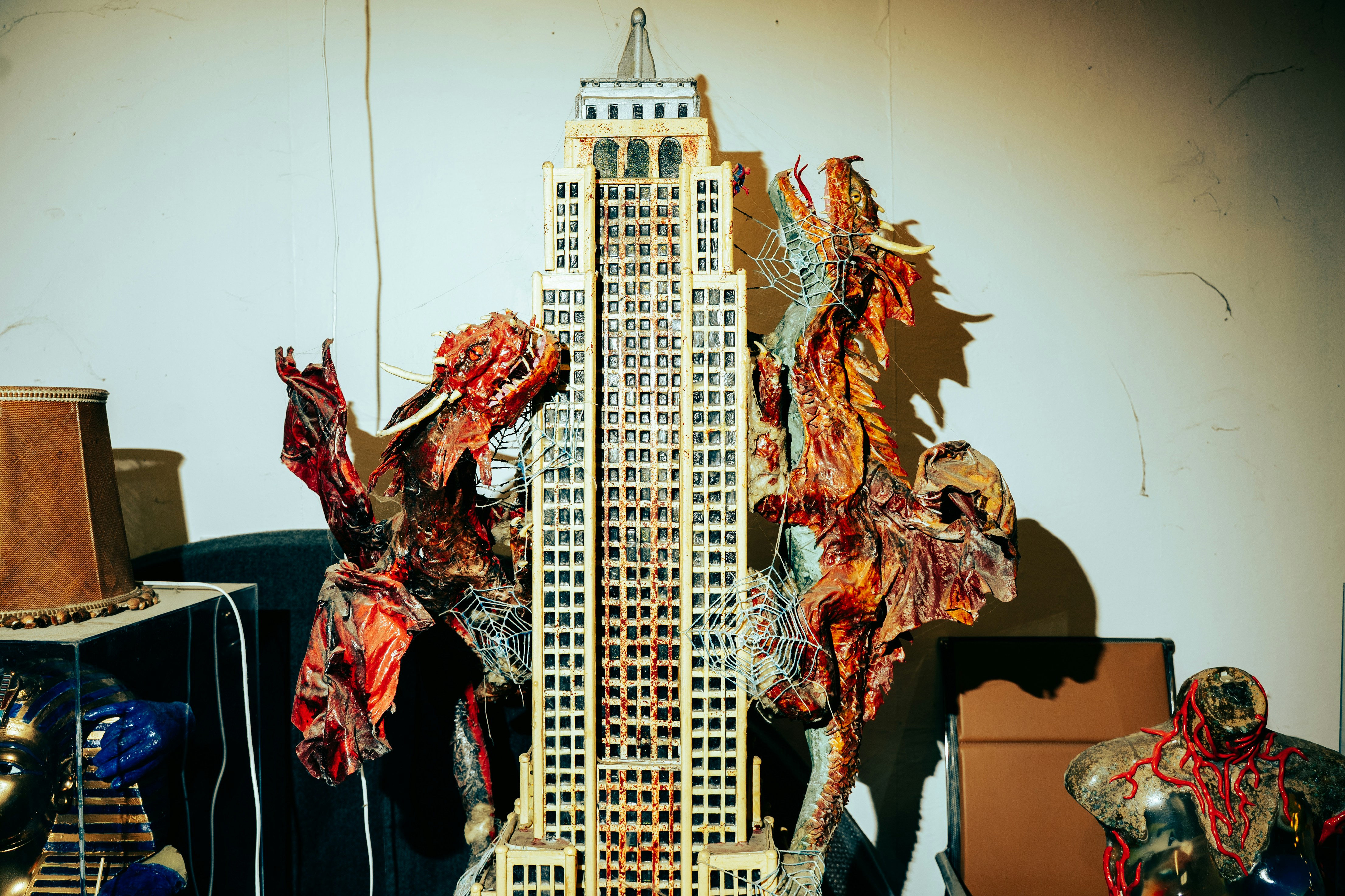 A tall stylized model tower sits center stage among chaotic toy figures and cluttered props in a cluttered indoor setup.