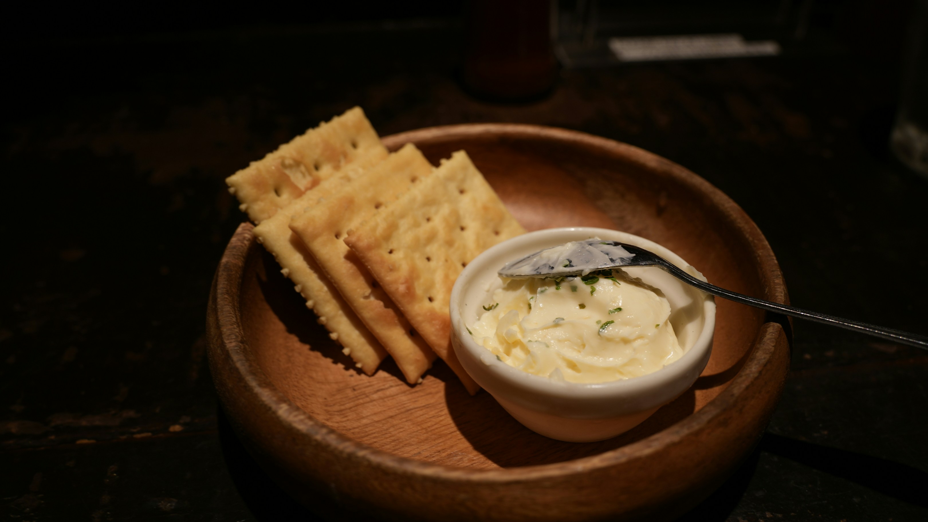 Cream cheese dip with herbs and a stack of crackers in a warm wooden bowl on a dark table.