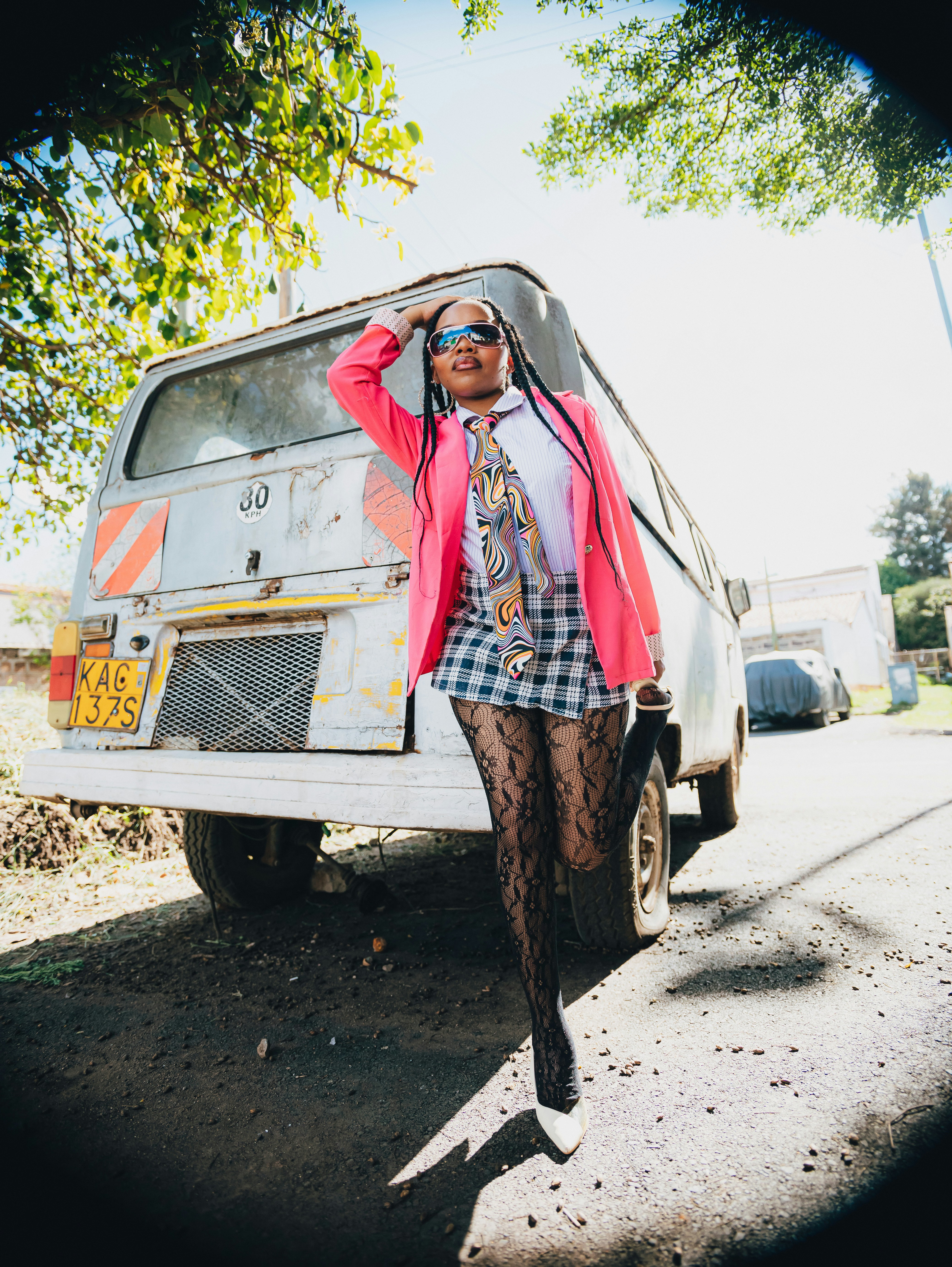 A fashion-forward individual stands confidently beside a weathered van, combining bold attire with a gritty outdoor setting. The scene emphasizes style against an urban backdrop.