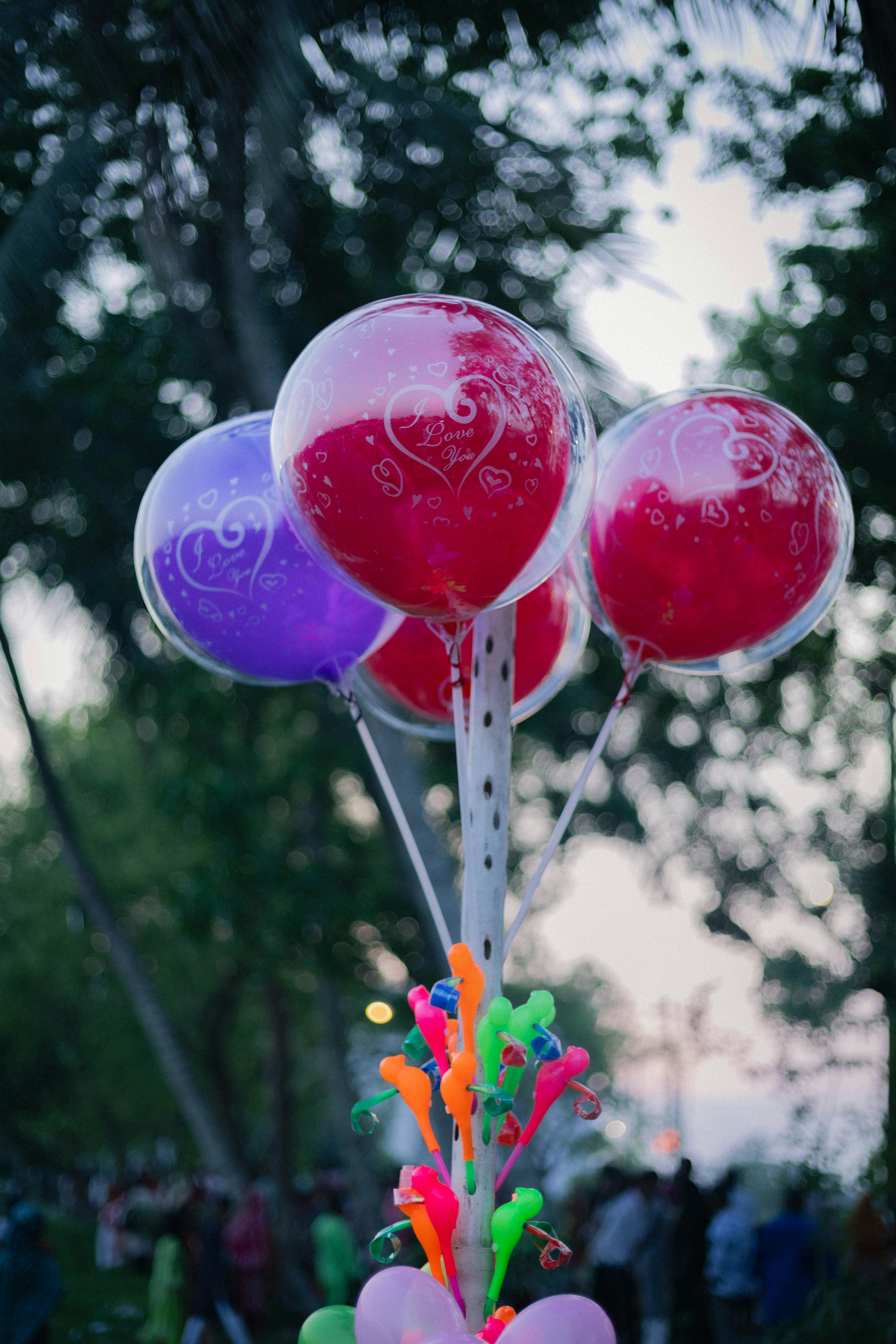 Colorful balloons rise above a festive stake in a park, with blurred trees and silhouettes in the background. A string of vibrant shapes anchors the composition.