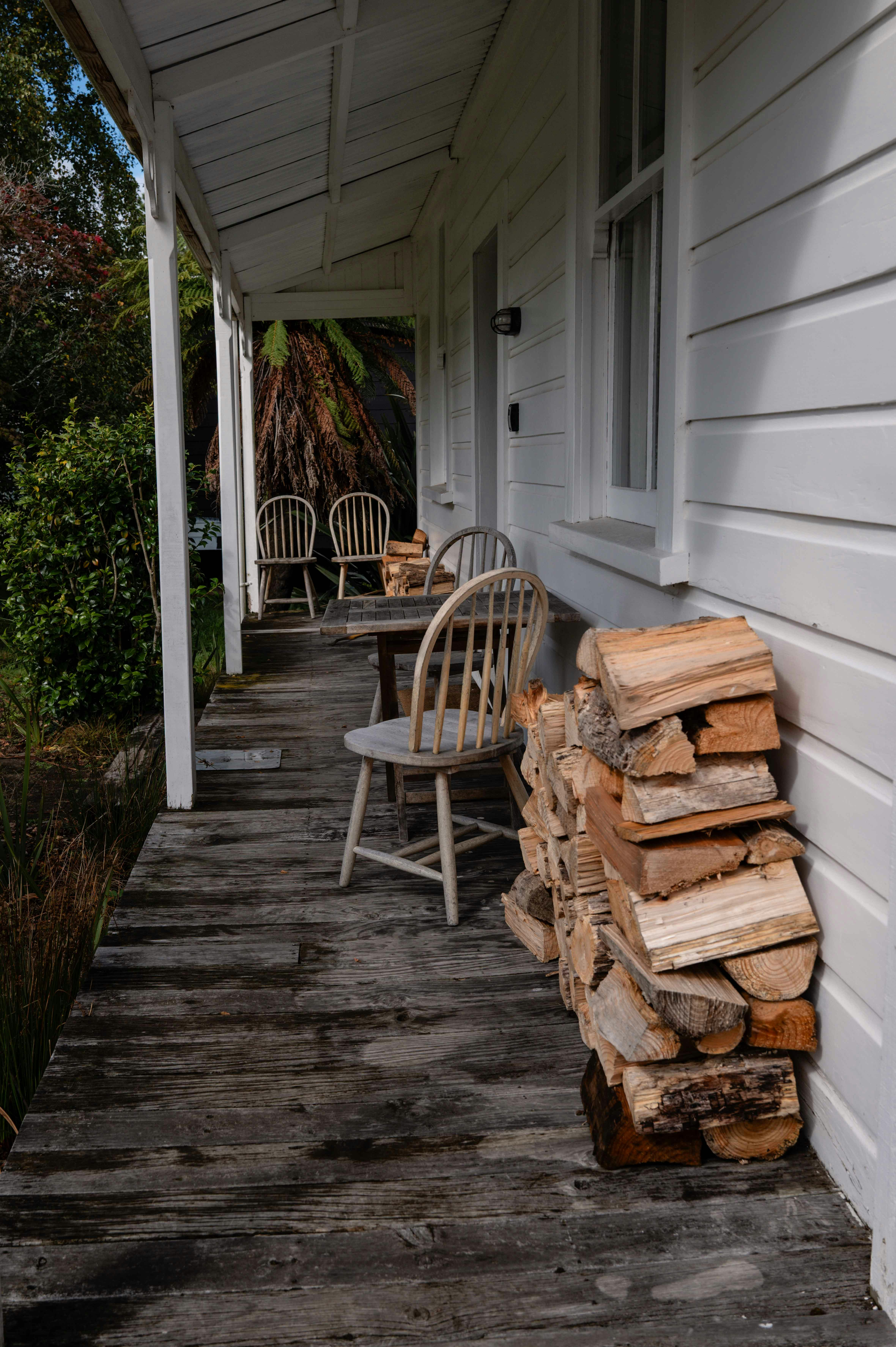 Stacked firewood and weathered porch along a sunlit white-washed house