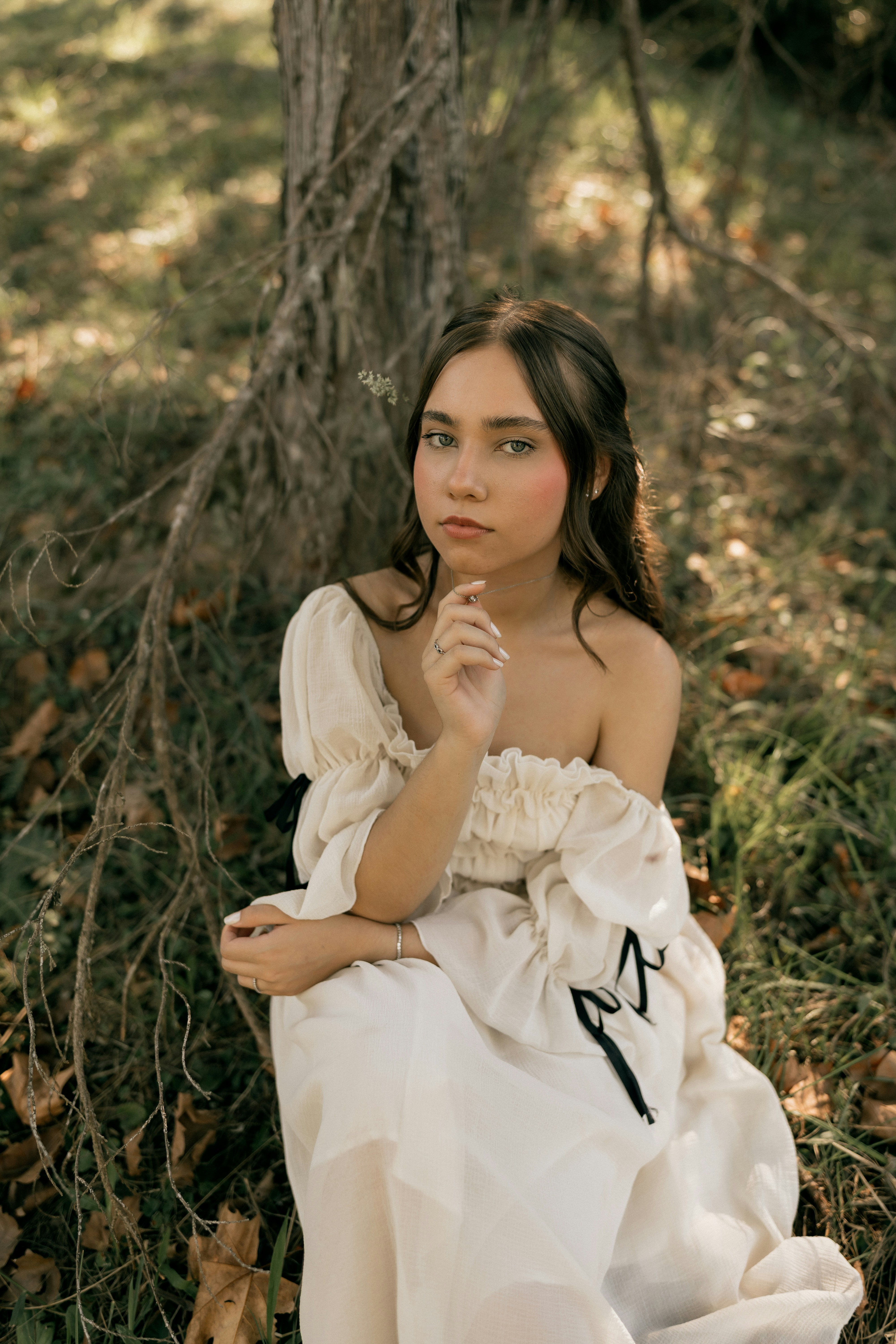 Young woman in a flowing dress sits by a tree.