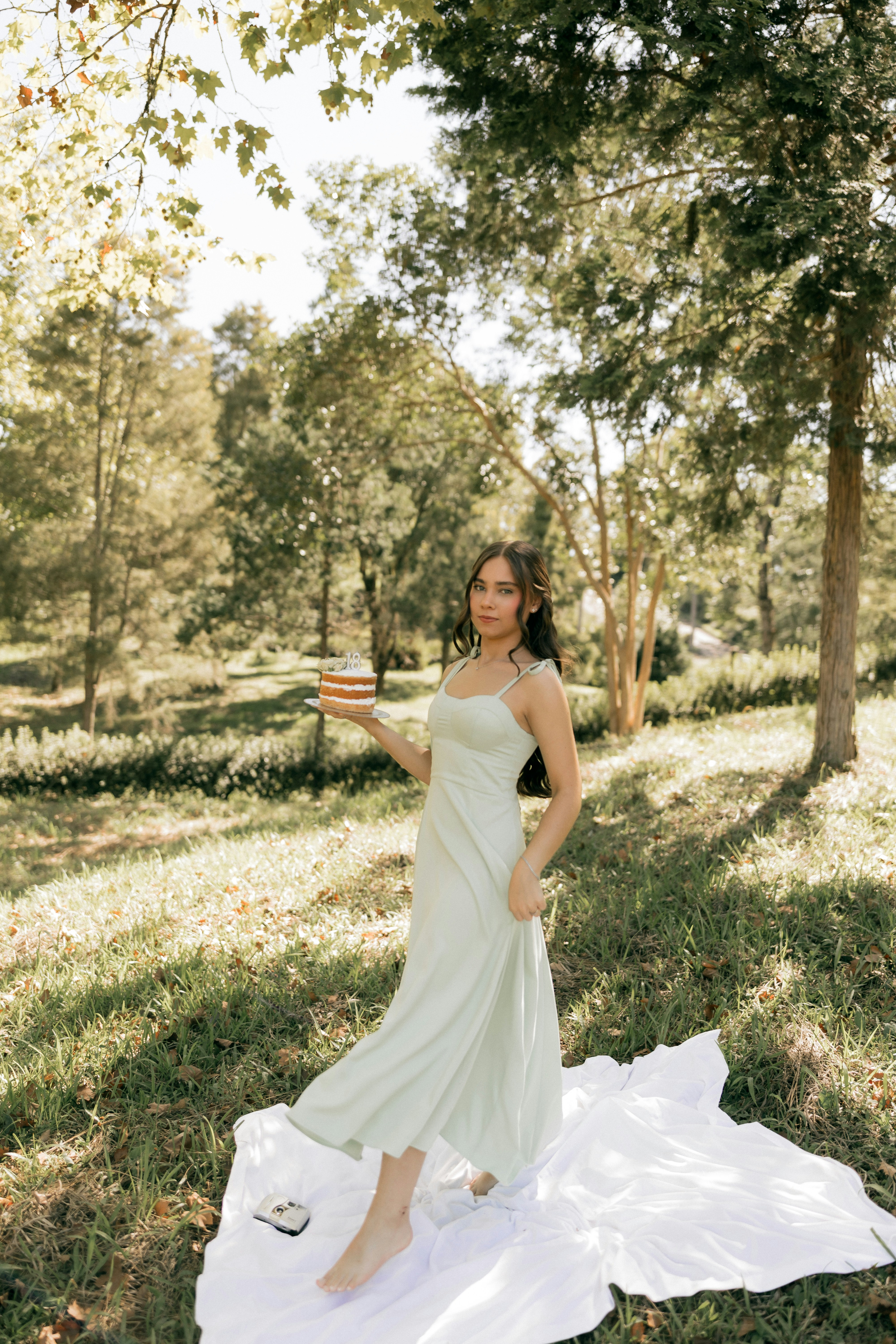 Bride-to-be in a pale-green dress holds a cake in sunlit park