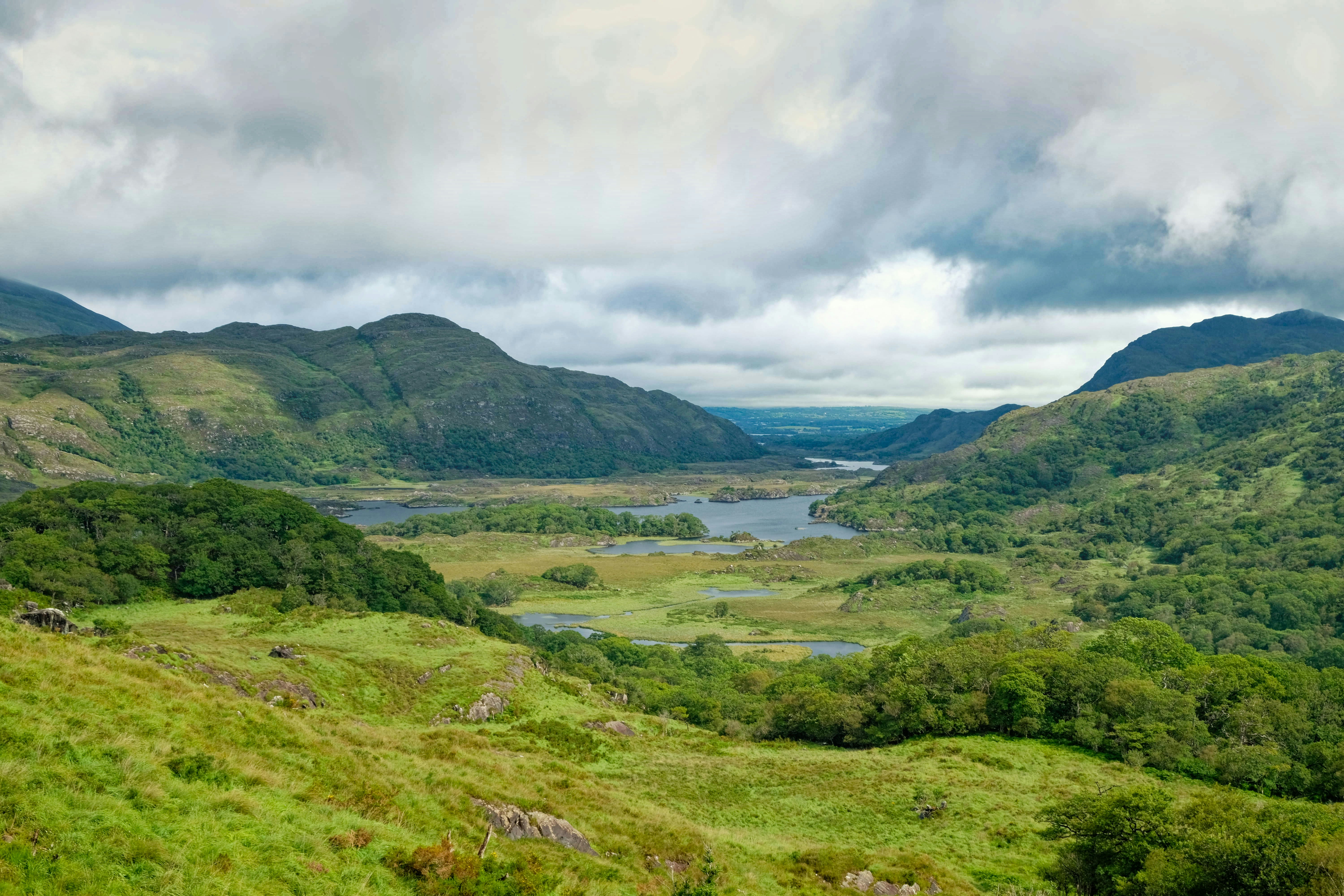 Lush valley stretches between rolling hills with a winding watercourse, under a cloudy sky.