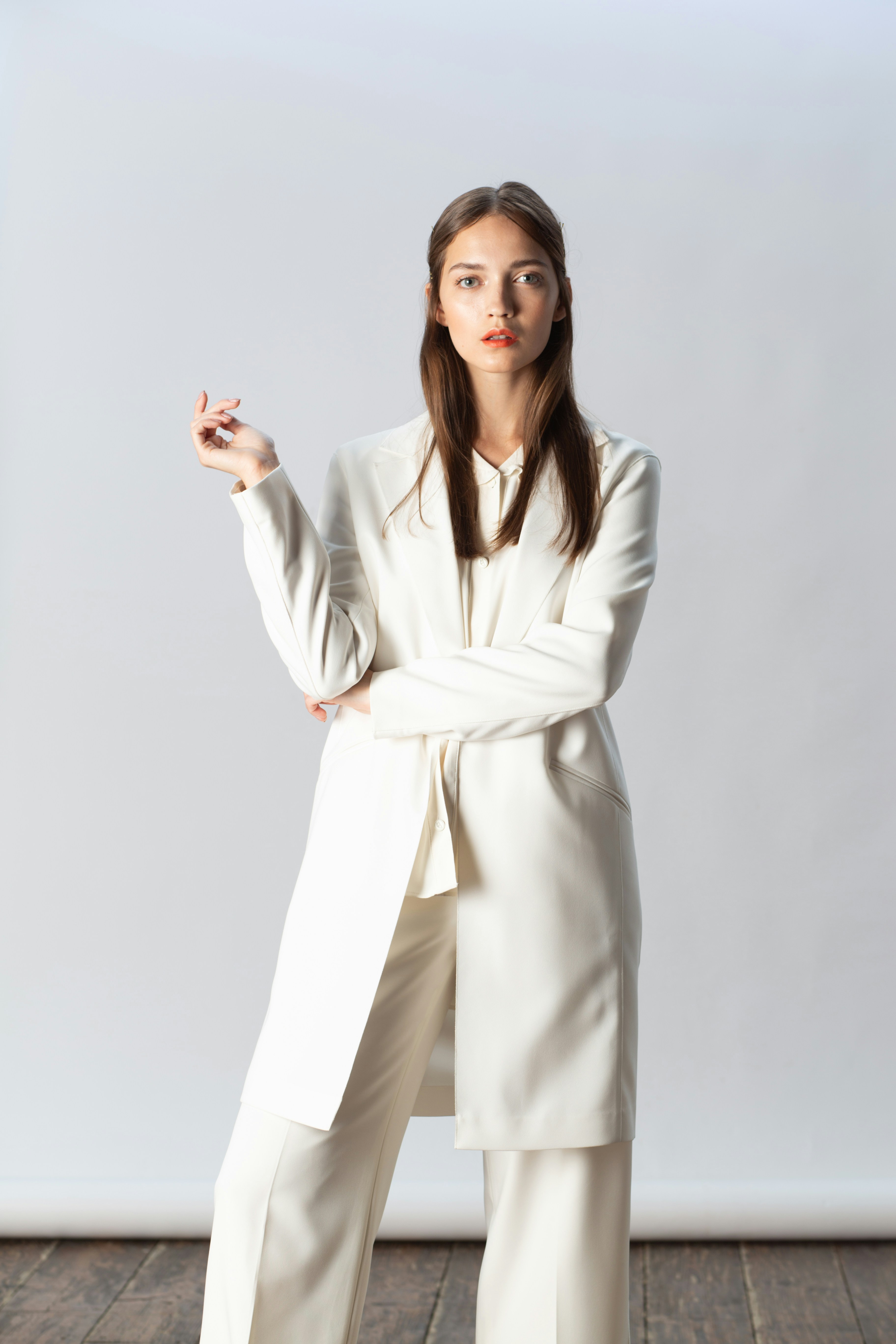 A poised woman in an ivory suit stands against a pale backdrop, arms crossed with one hand lightly lifted. The clean studio setup emphasizes minimalism and refined style.
