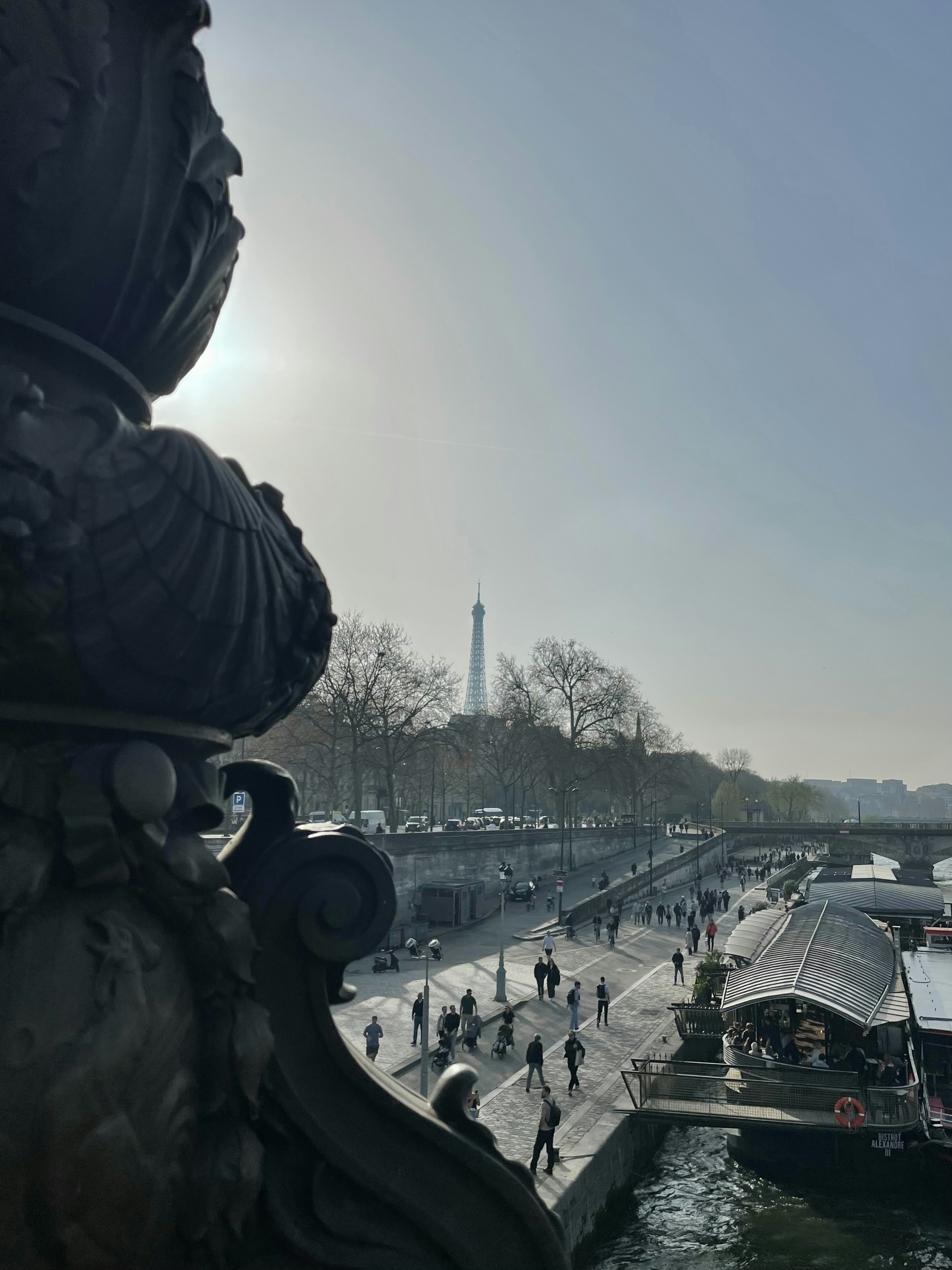 Sunlit riverside scene along the Seine with a decorative foreground balustrade and a boat docked near the bank; pedestrians populate the promenade while the Eiffel Tower rises in the distance.