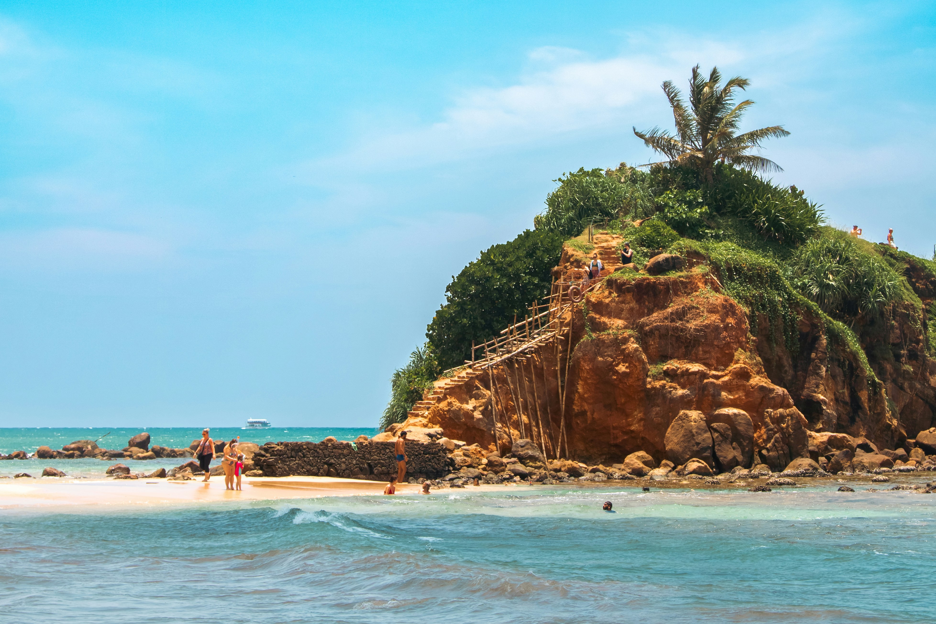 Parrot Rock in Mirissa, Sri Lanka, framed against a wide blue sky, with gentle waves in the foreground. Located on Mirissa Beach, Parrot Rock is a small rocky island set across shallow turquoise water just off the golden shore. Reaching it requires quick timing as waves roll in, adding a sense of adventure to the crossing. A narrow path leads to the top, offering sweeping views of the coastline and ocean. From the summit, you can spot Coconut Tree Hill in the distance.
