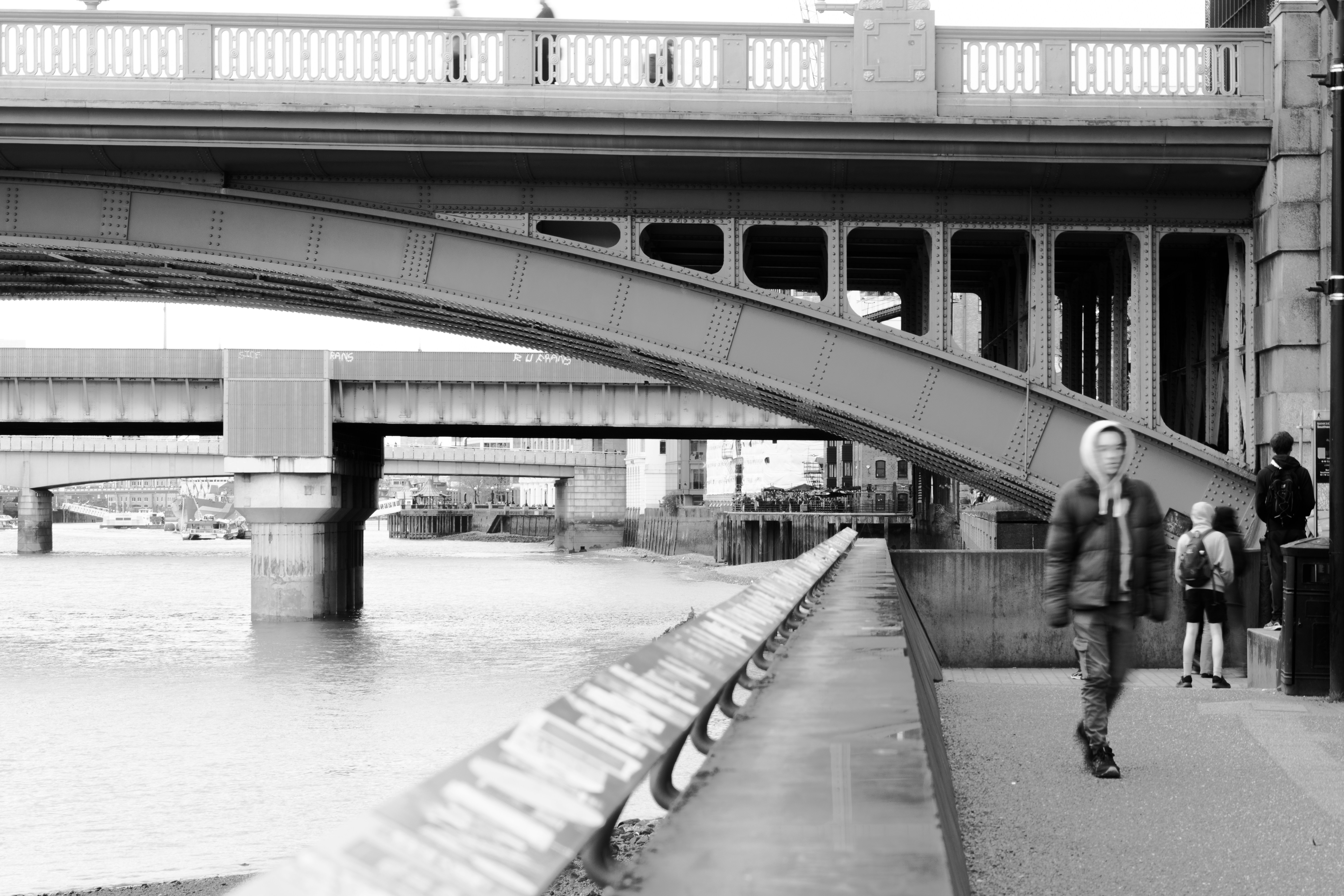 People walk along a waterfront beneath a large bridge.